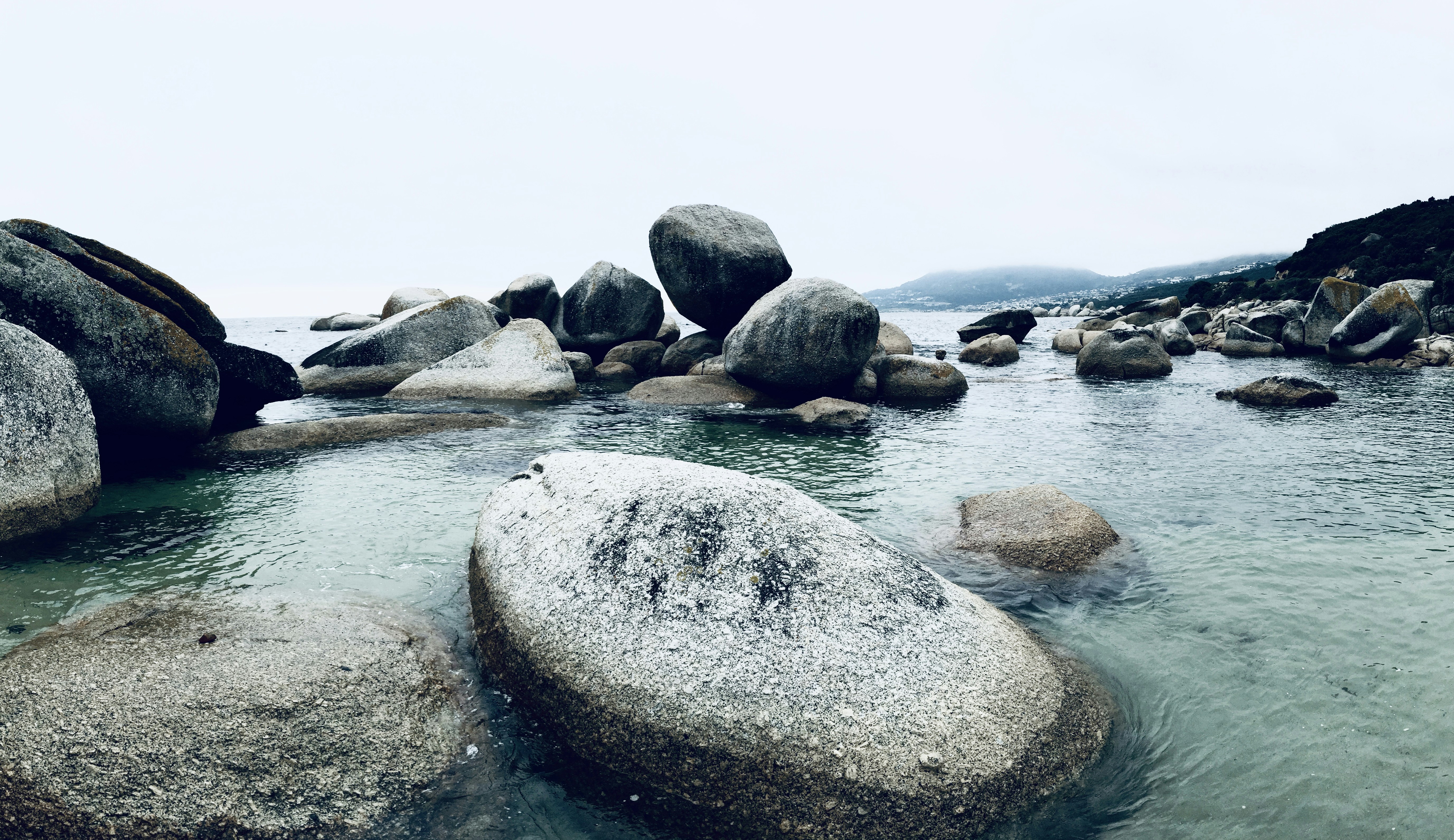 Smooth, weathered boulders emerge from calm waters, reflecting a muted sky and distant hills. The serene coastal landscape invites contemplation.