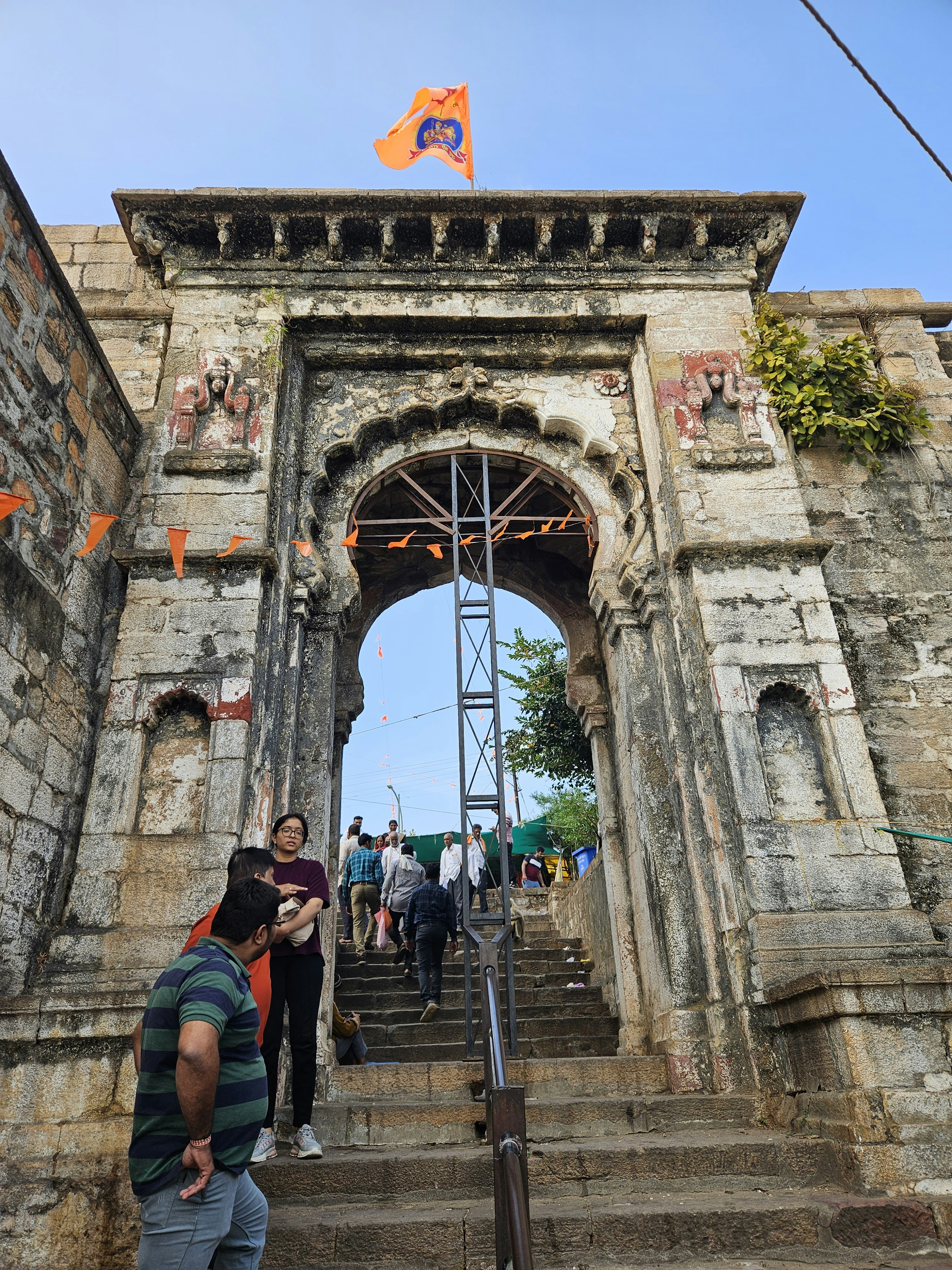 Rugged stone archway and weathered entrance framed by people ascending a stone staircase; flags and a line of visitors hint at a historic site or fortress, with a bright blue sky above.