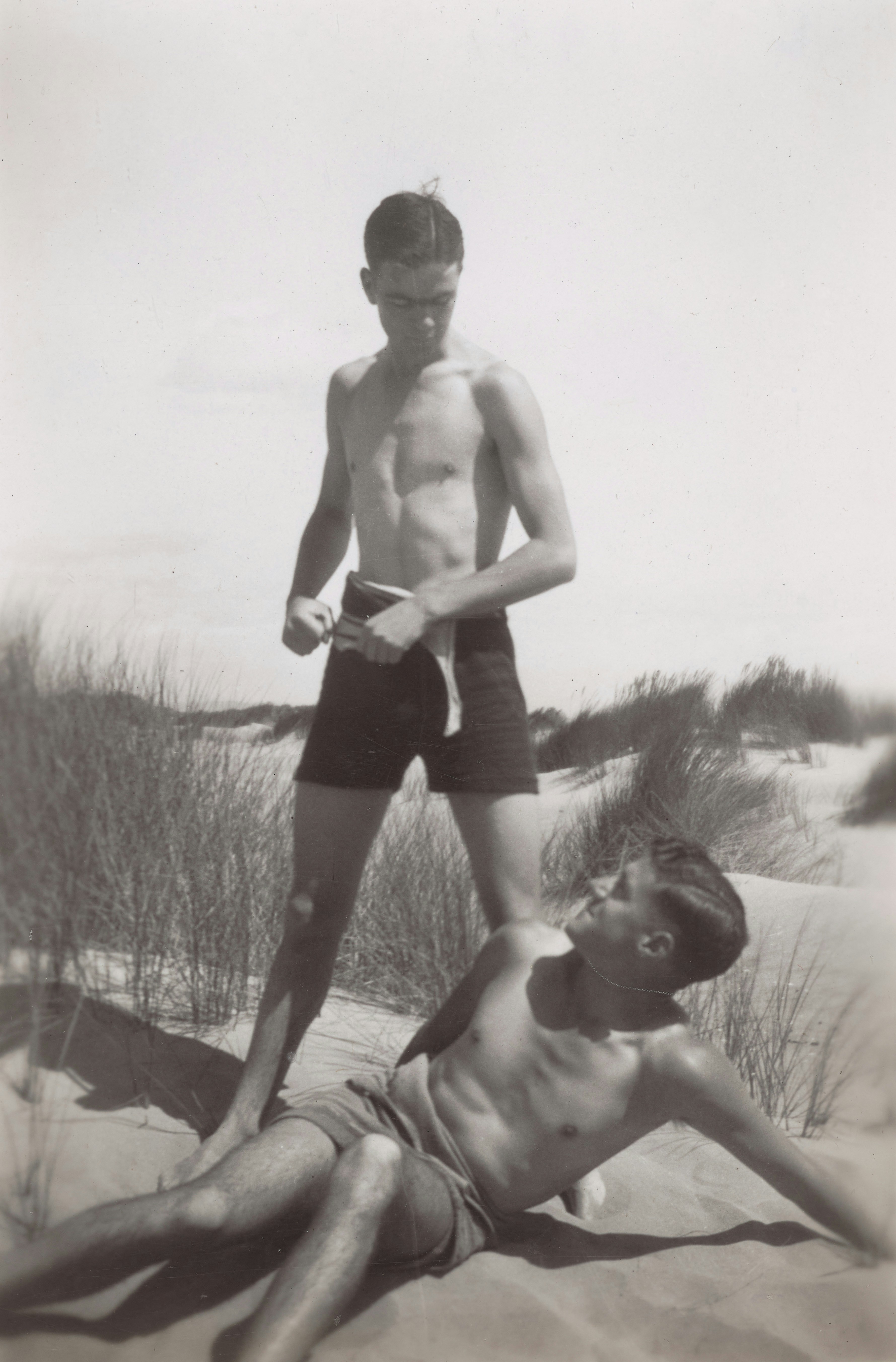 Two shirtless men on a windswept dune—one standing and adjusting his belt, the other reclining in the sand—captured in a high-contrast black-and-white photograph.
