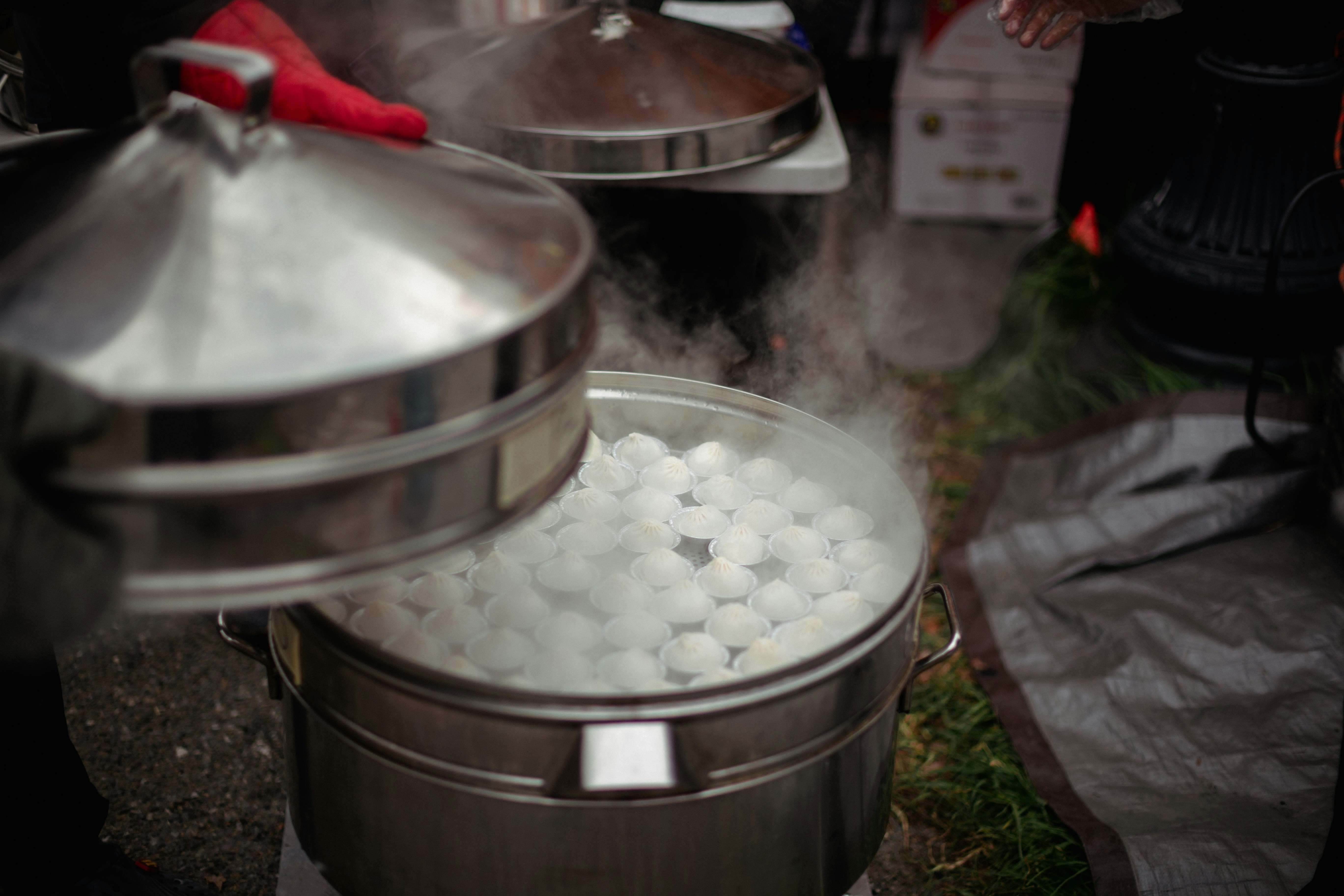 Steaming bamboo-dasket pot with a lid ajar reveals round dumplings rising above steam in an outdoor setup.