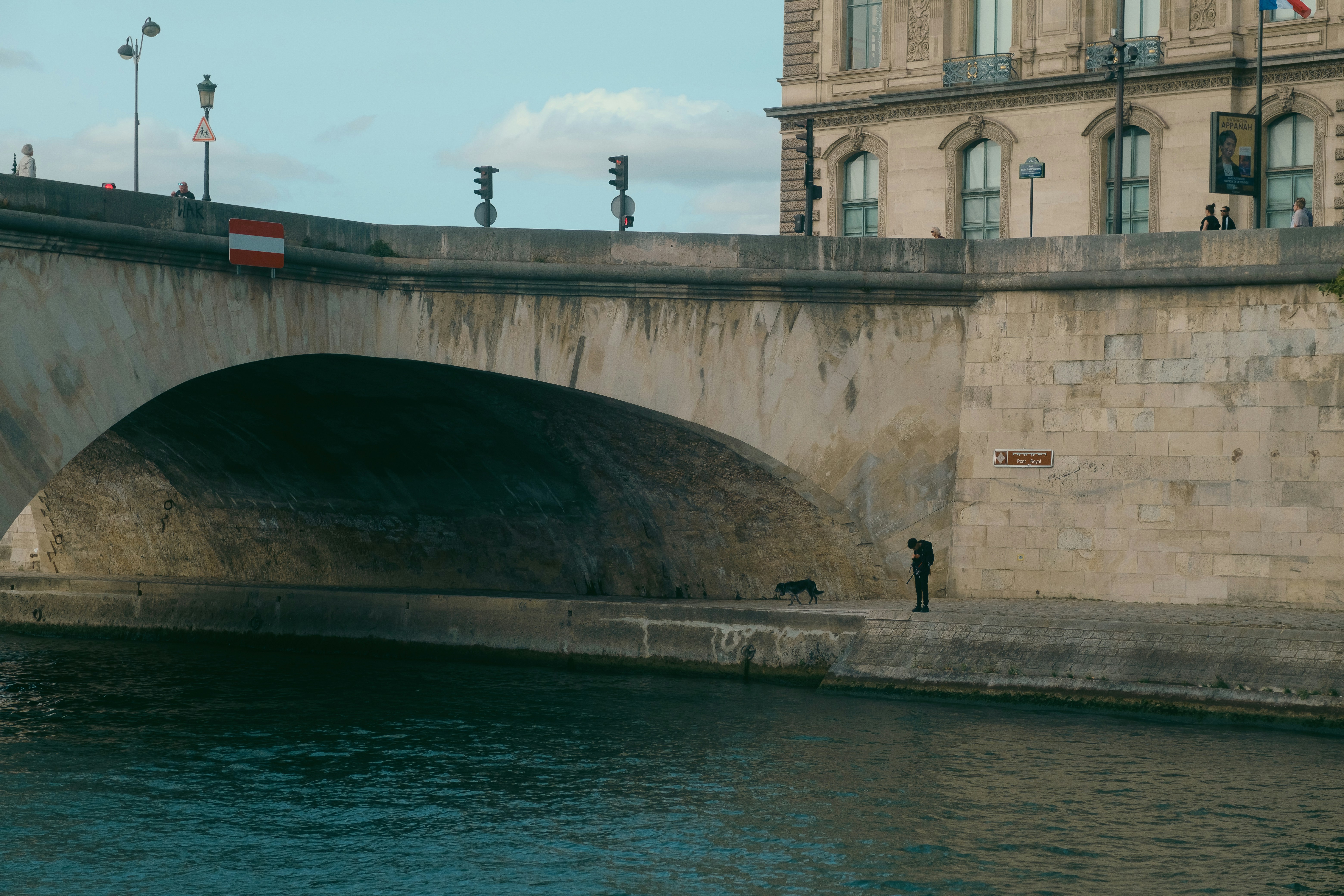 Person with an umbrella walking beneath a historic stone bridge by a river.