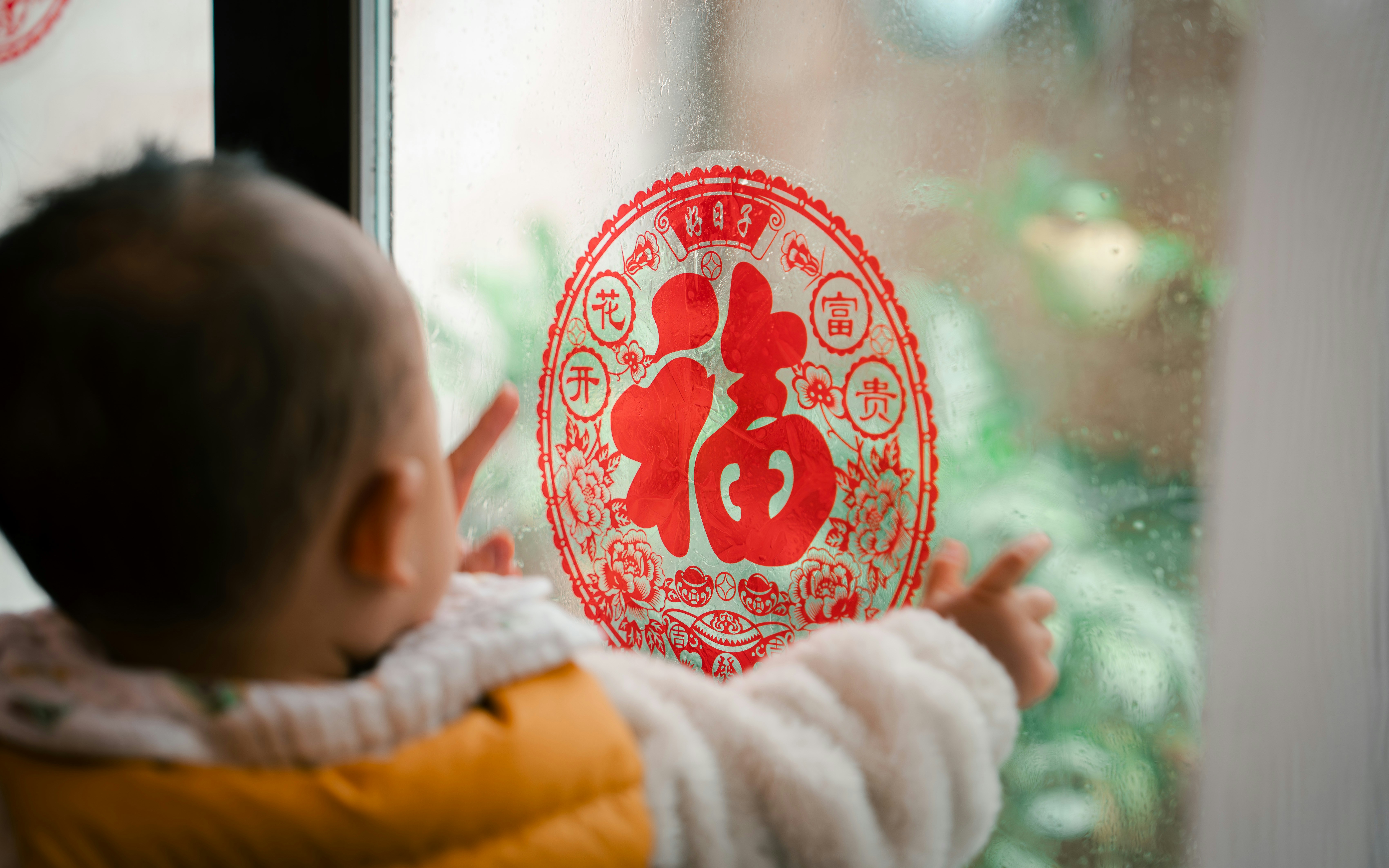 Child reaching out to touch a red paper decoration on a foggy window, symbolizing good fortune. The soft focus creates a warm, nostalgic atmosphere.