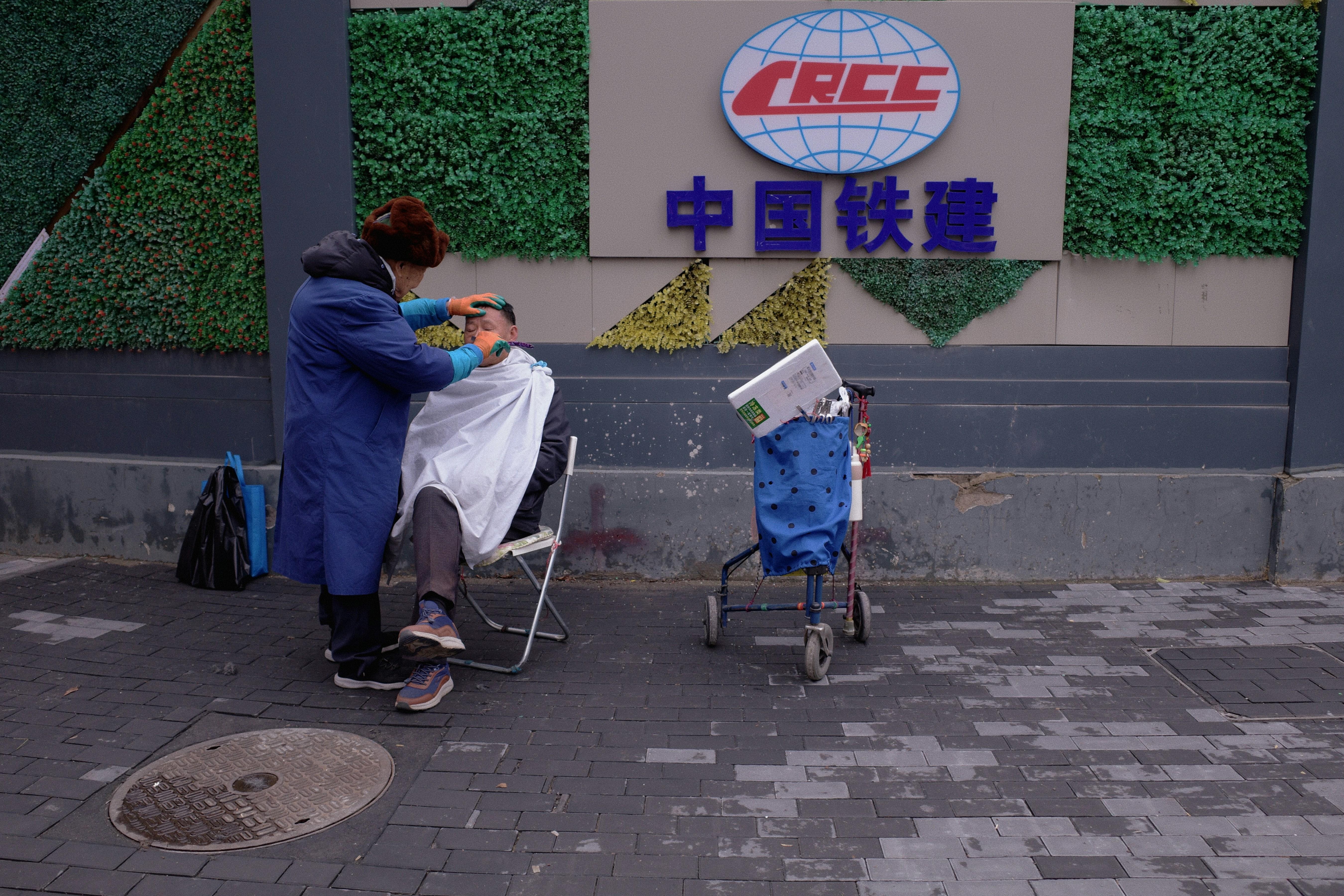 Photograph of a street-side barber trimming a client on a sidewalk, with a storefront sign and a wheeled cart against a green ivy-covered wall.
