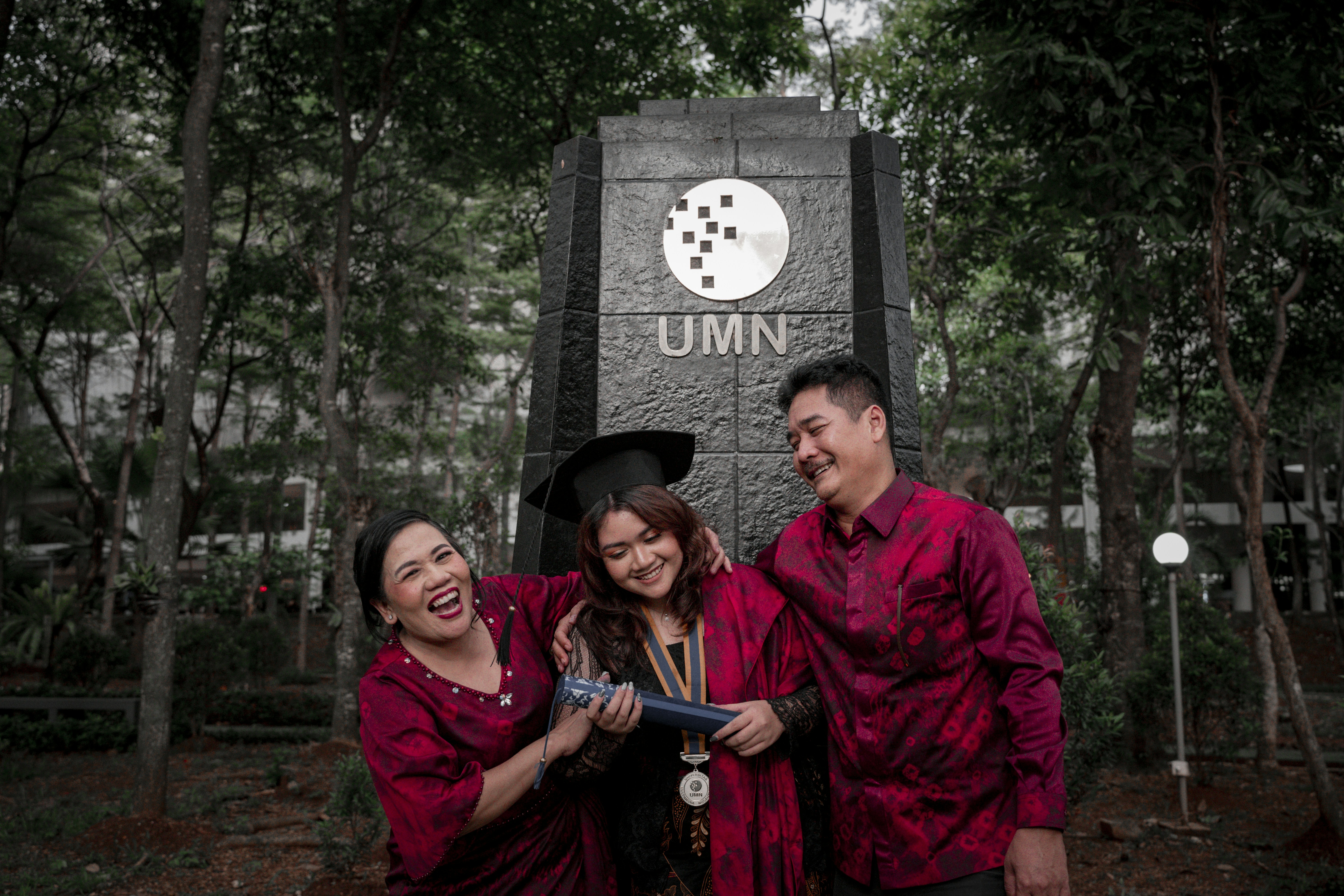Three people in red attire share a joyous moment in front of a UMN sign amidst trees.