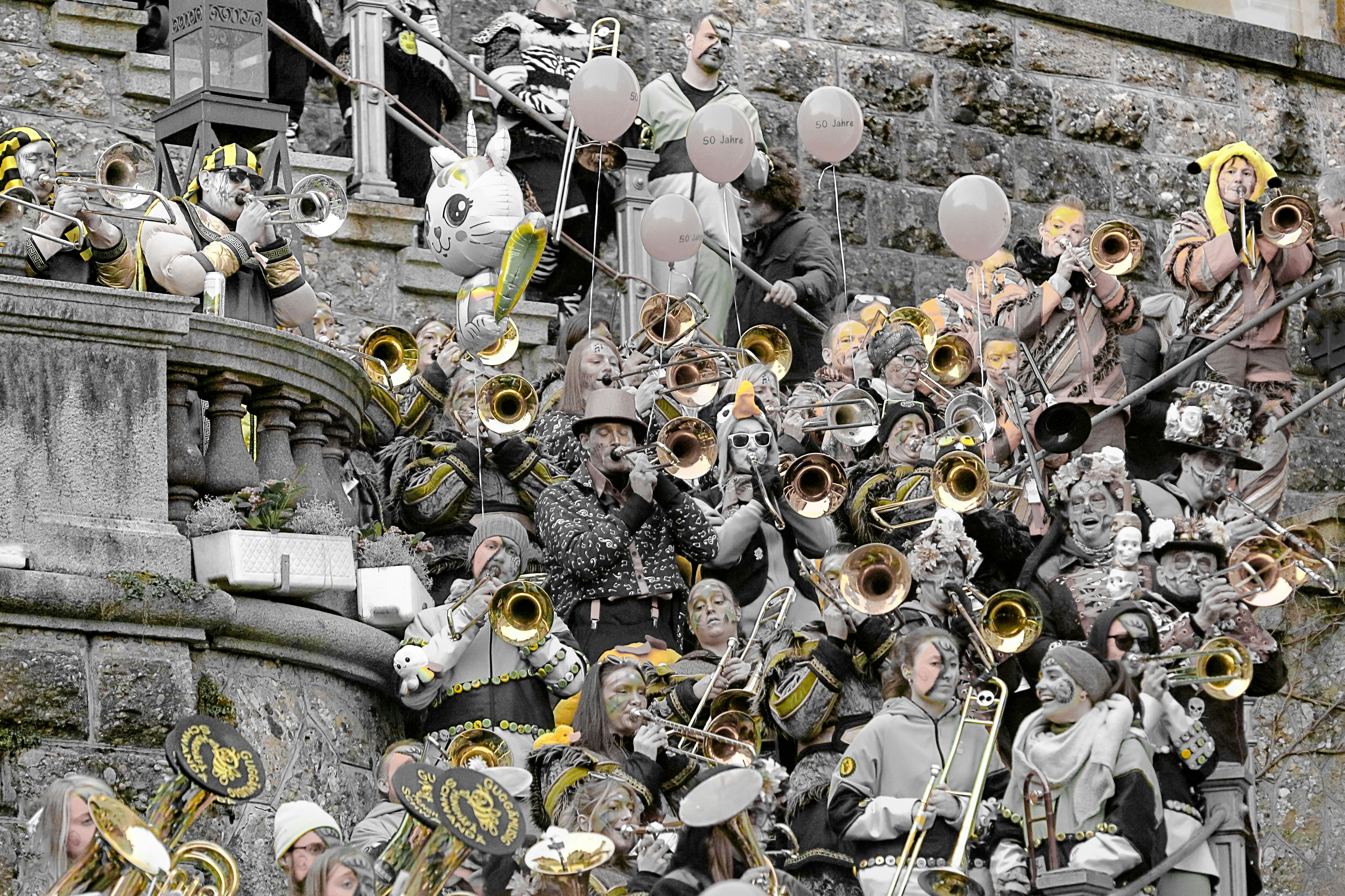 Large group of musicians playing brass instruments on stone steps, holding balloons amid historic architecture.