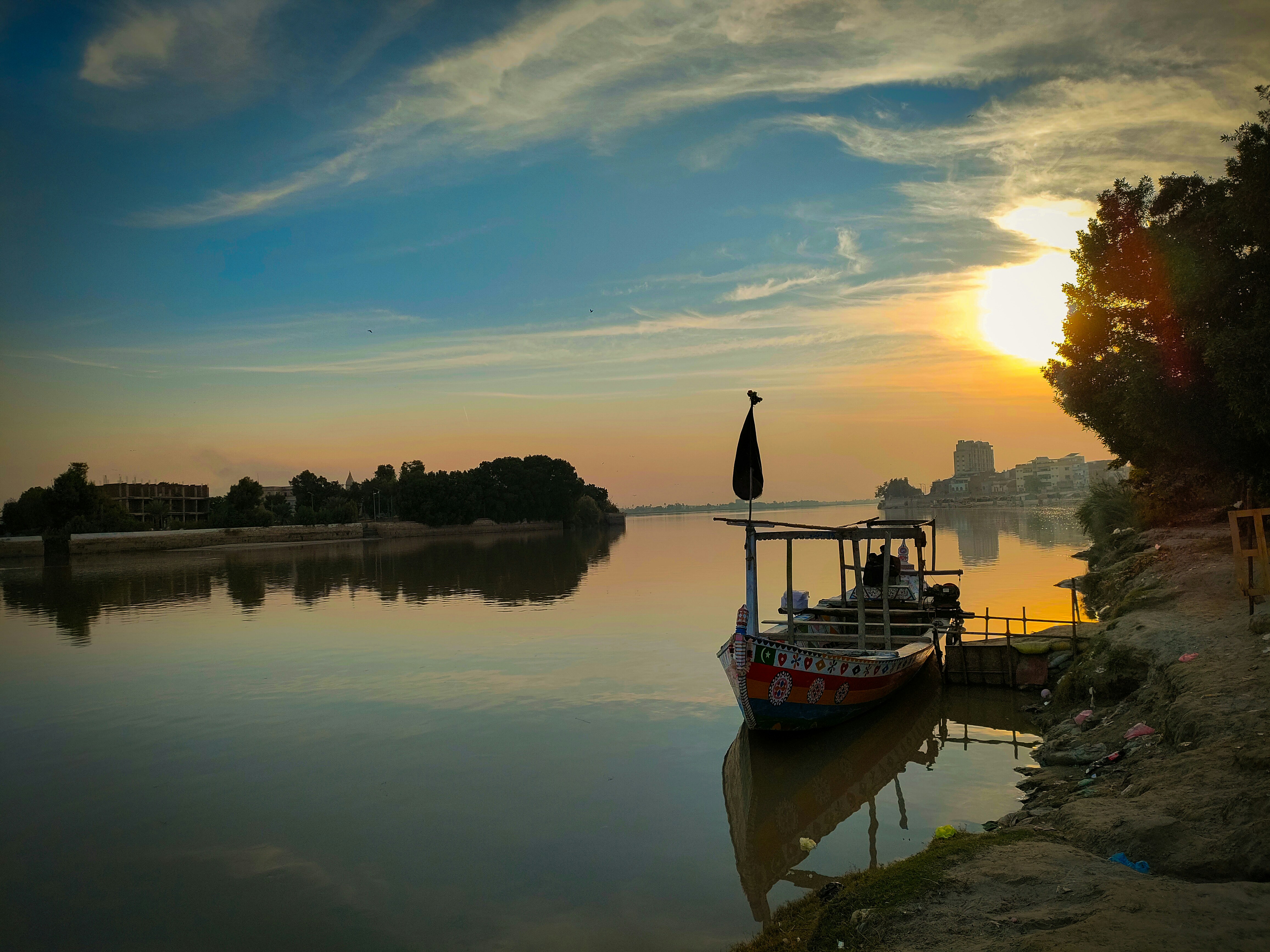 Boat docked on a calm river at sunrise with silhouetted trees and buildings in the background.