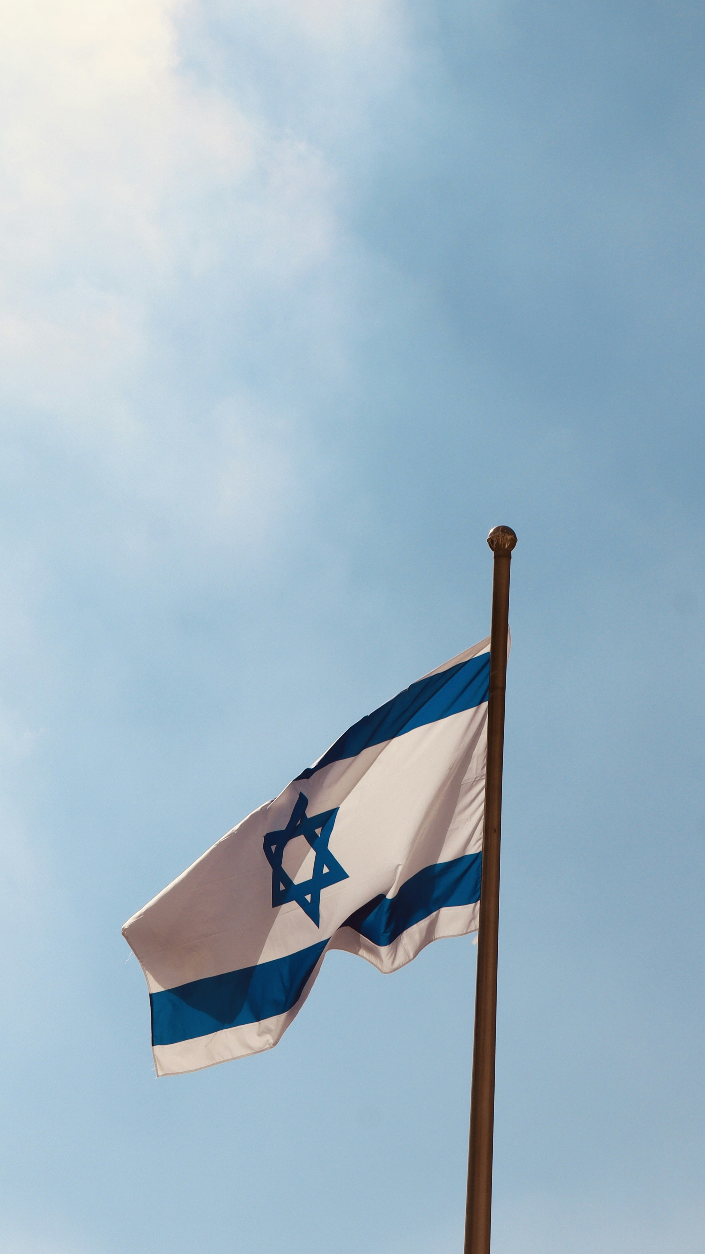Israeli flag billowing in the breeze, set against a backdrop of soft blue skies. The design features blue stripes and a Star of David.