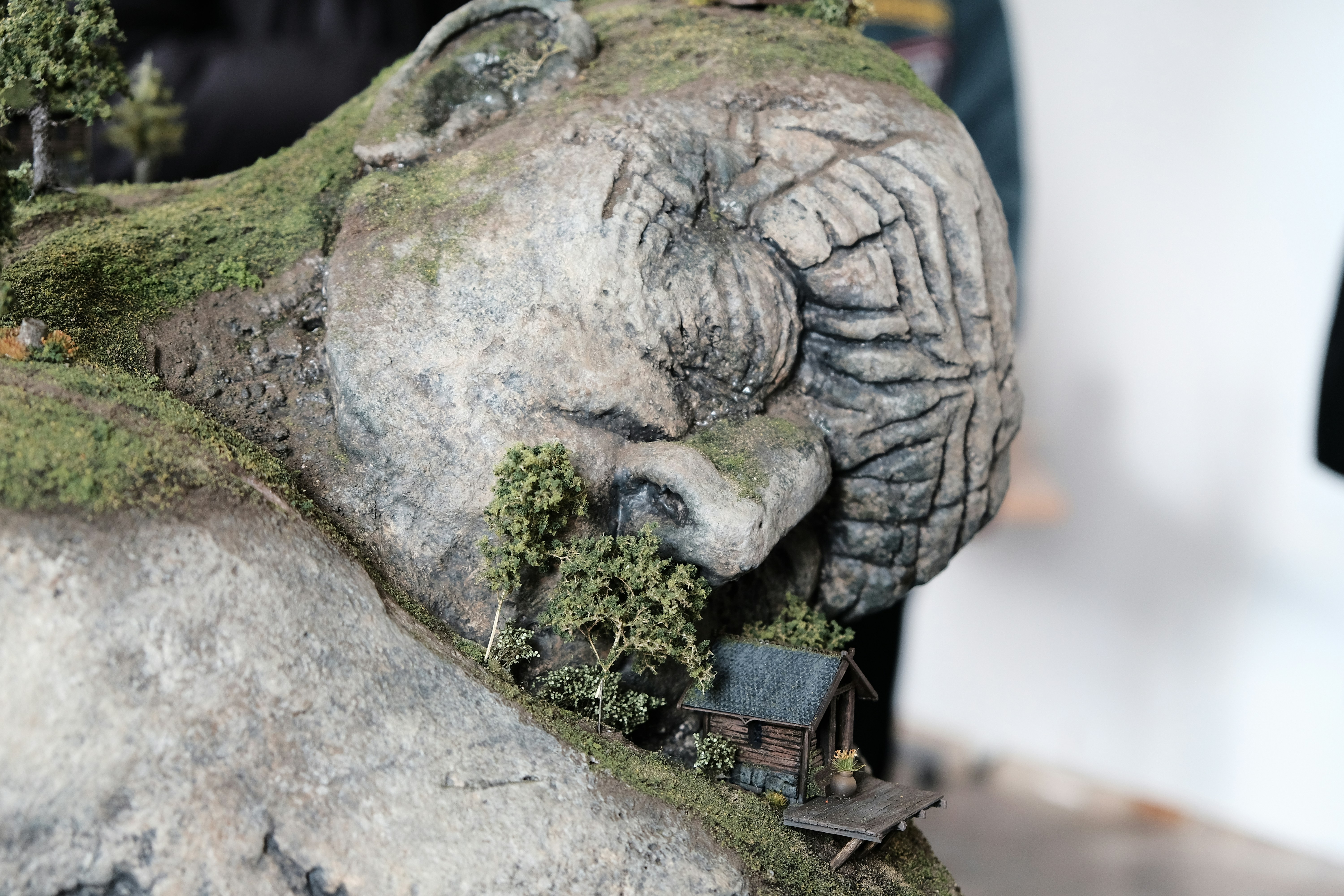 Concrete sculpture of a sleeping giant with moss and a tiny house nestled in its folds.