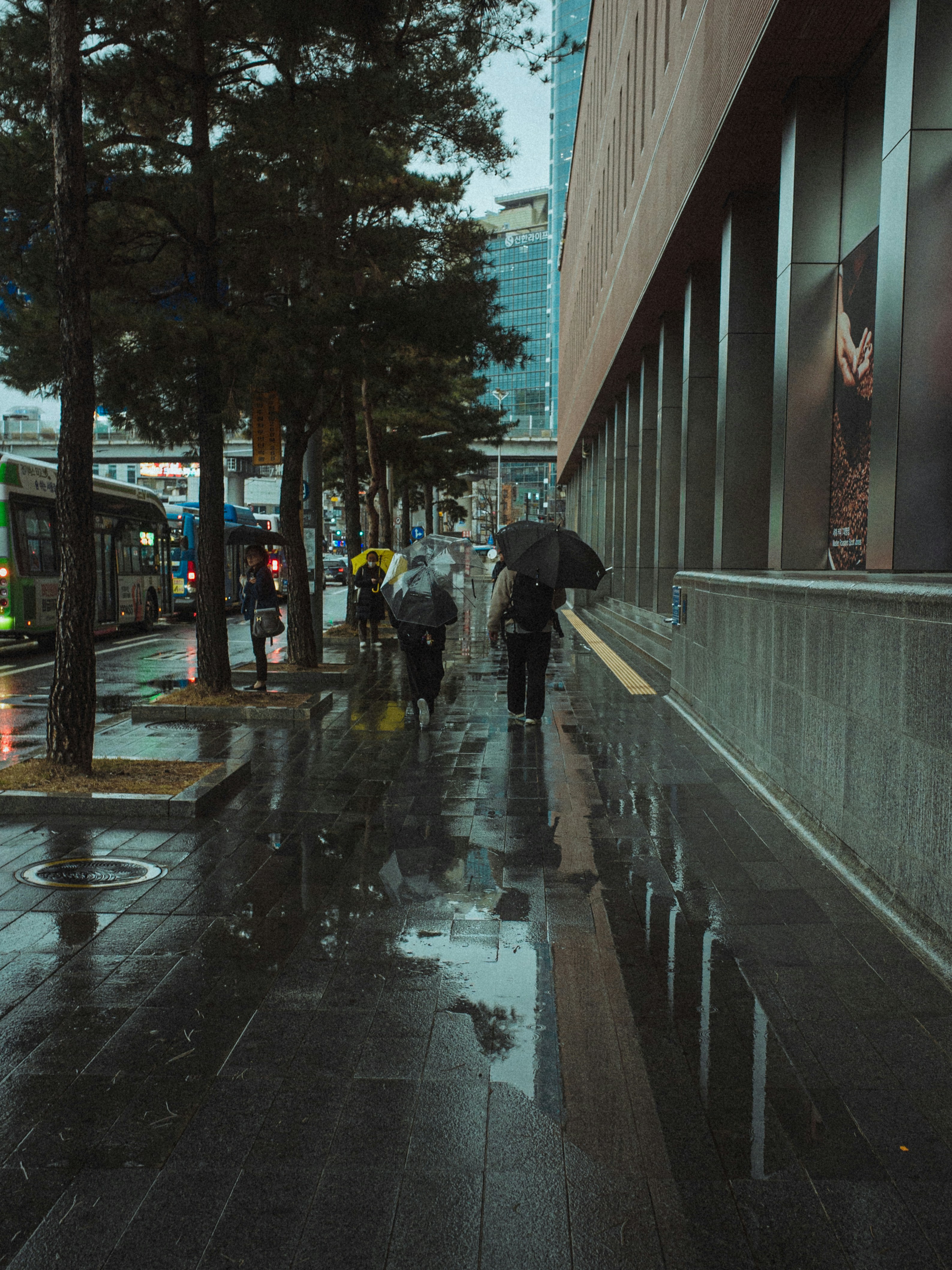 Rain-soaked city sidewalk with pedestrians under umbrellas. Wet reflections shimmer along the curb beside a modern building.