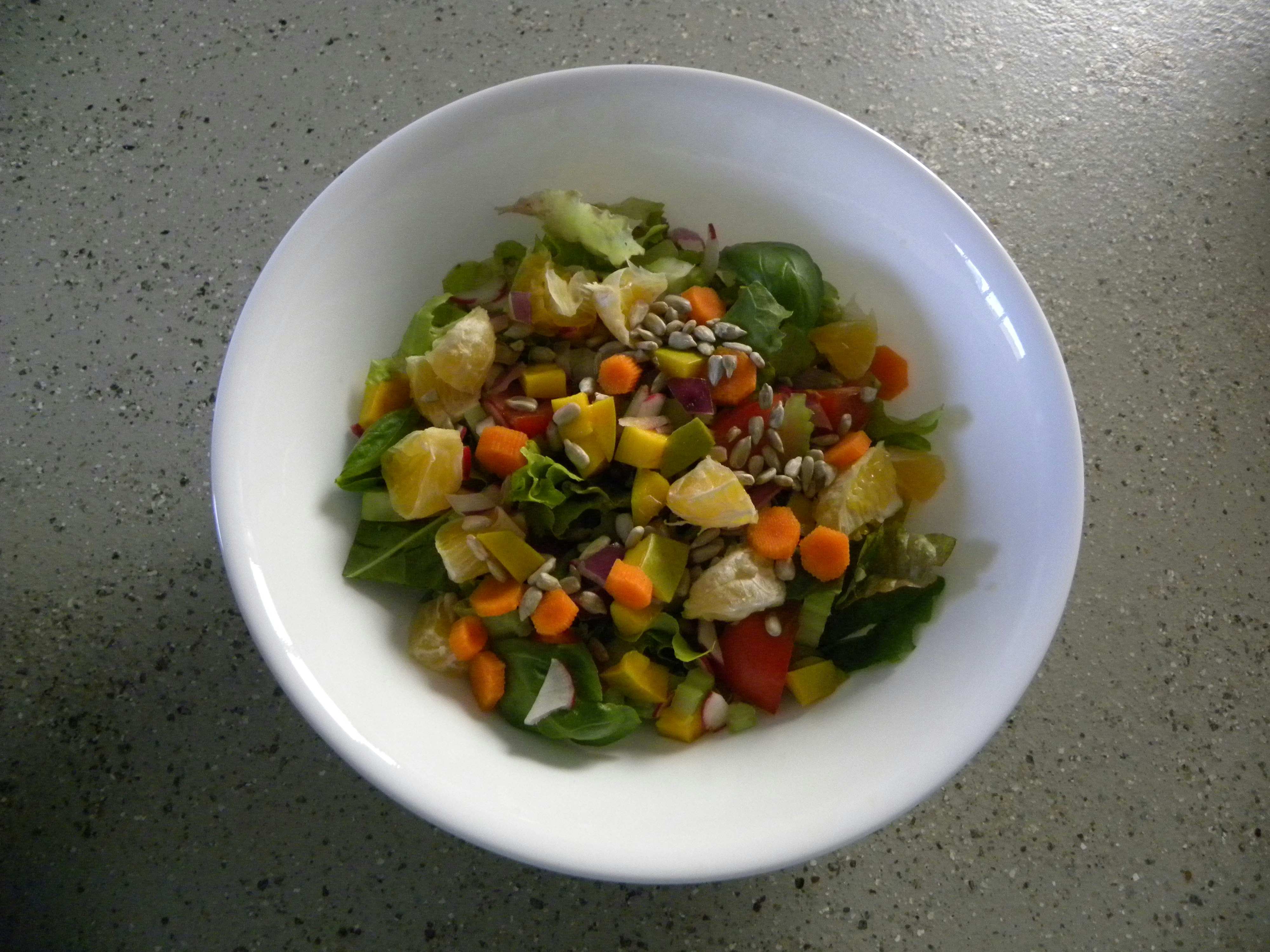 Colorful chopped vegetables and seeds in a white bowl sit on a gray speckled countertop. The composition highlights freshness and texture.