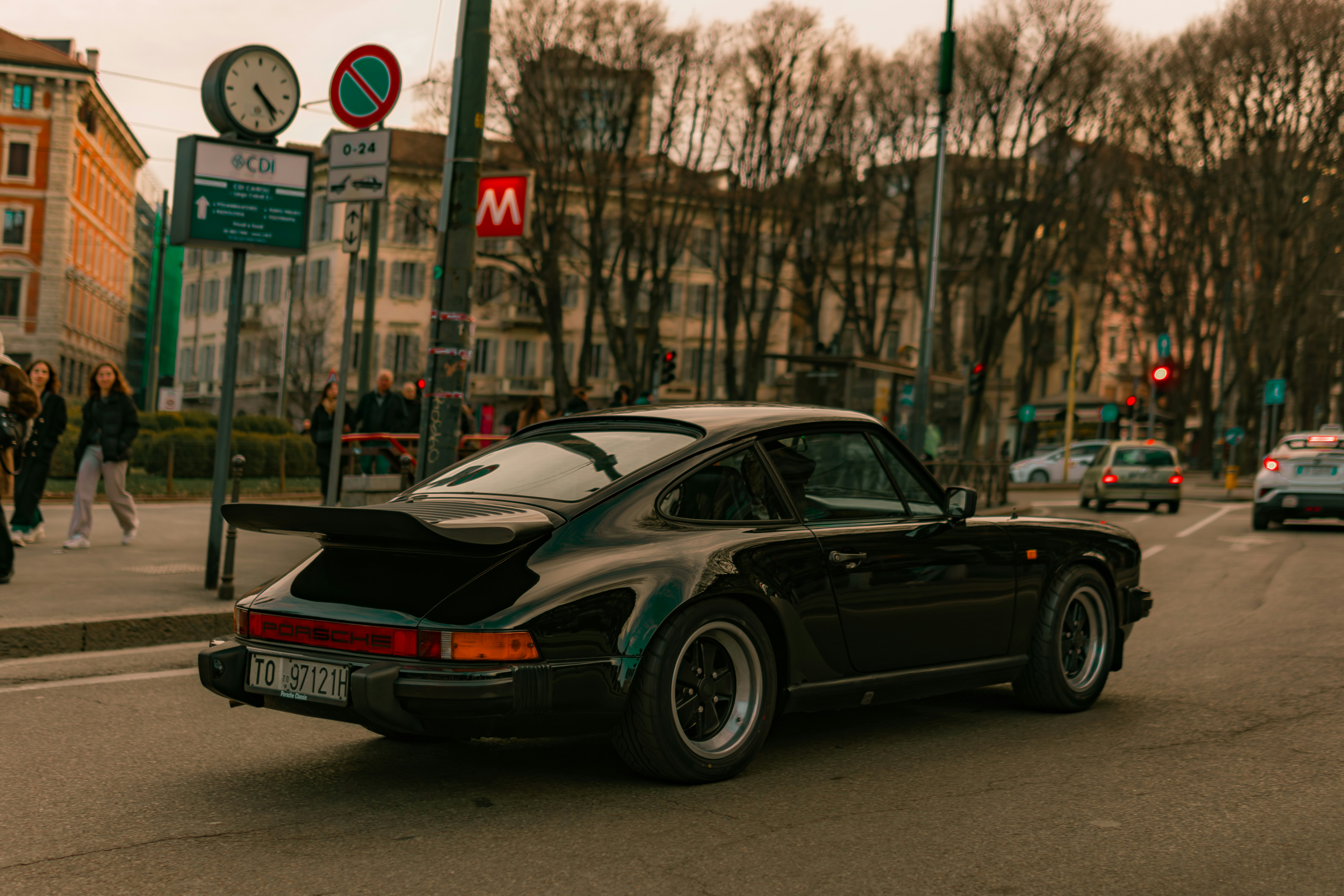 Photograph of a black Porsche 911 on an urban street with pedestrians and city signage in the background.