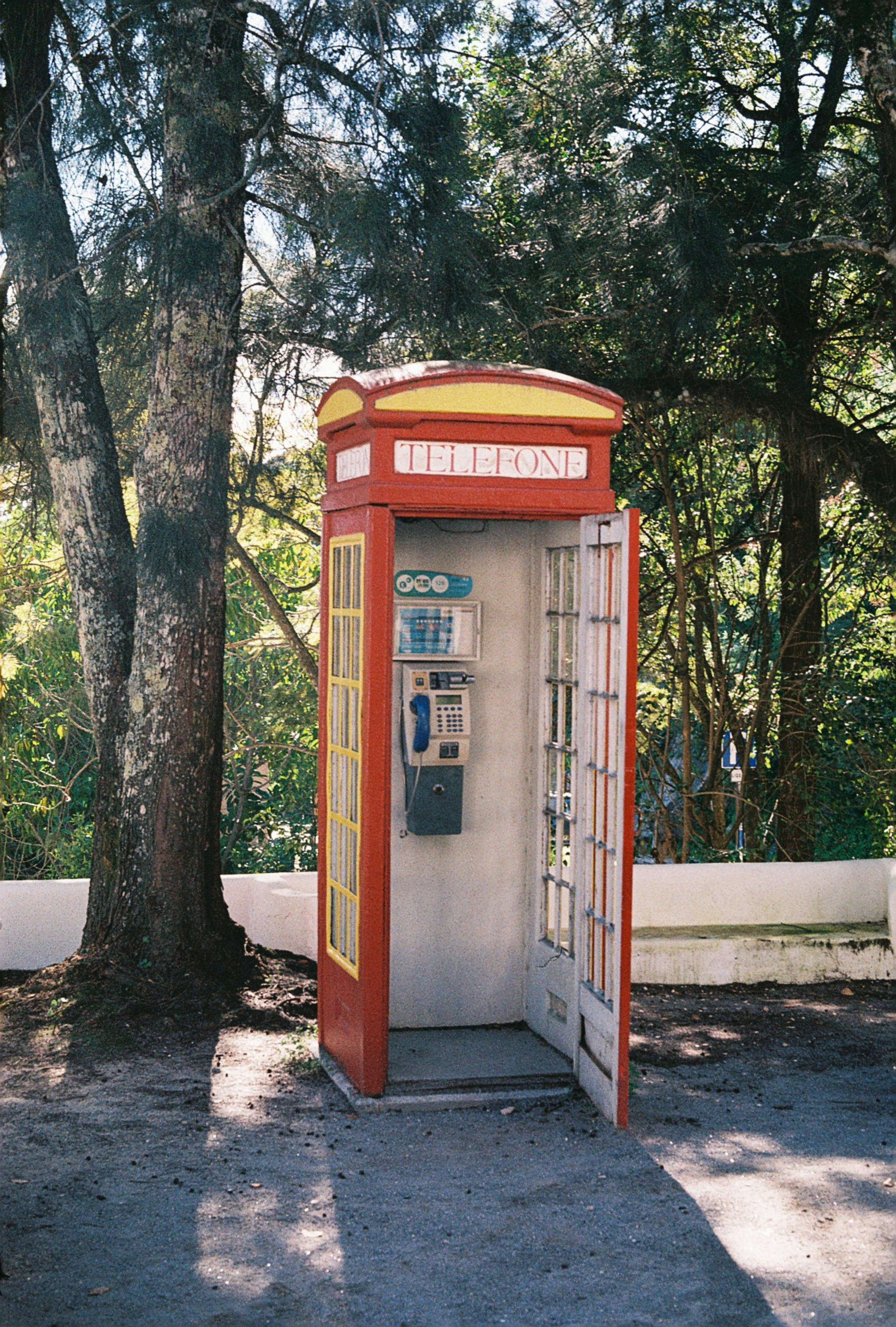 A classic red telephone booth stands beside a tree-lined path in a sunlit forest. The open door reveals the weathered interior and vintage hardware.