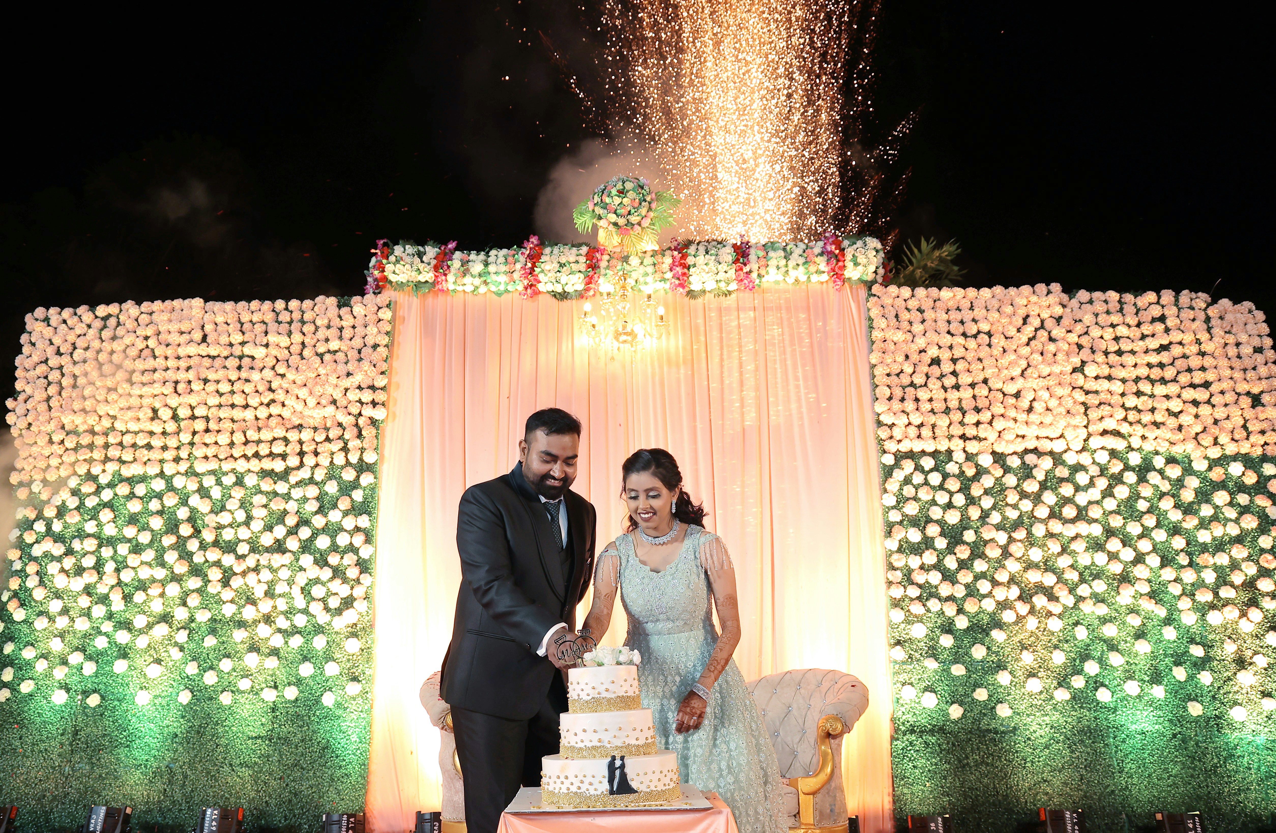 Couple cutting a tiered cake in front of a floral backdrop with fireworks overhead.