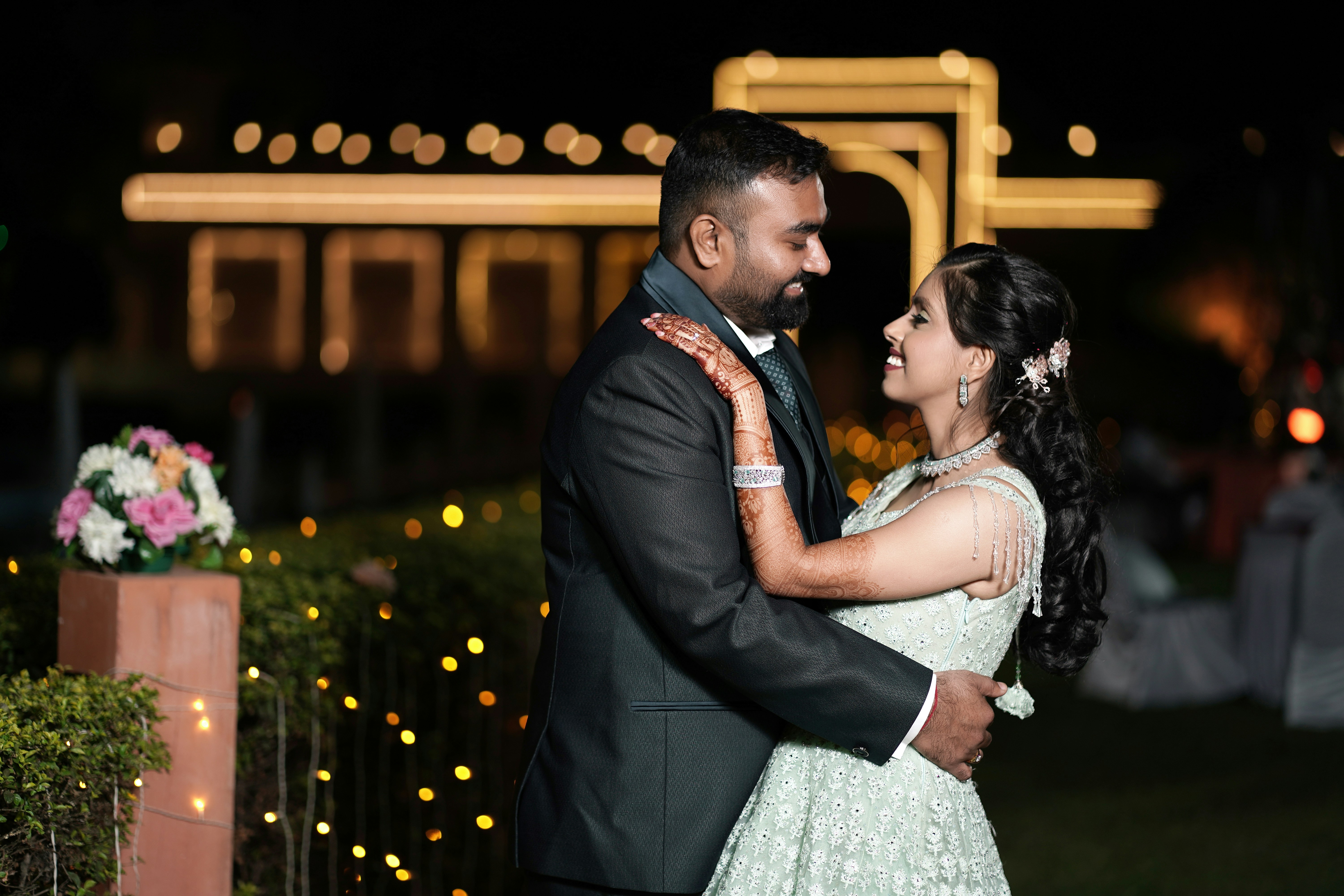 Couple shares a joyful gaze in an evening garden setting with twinkling lights and a floral arrangement.
