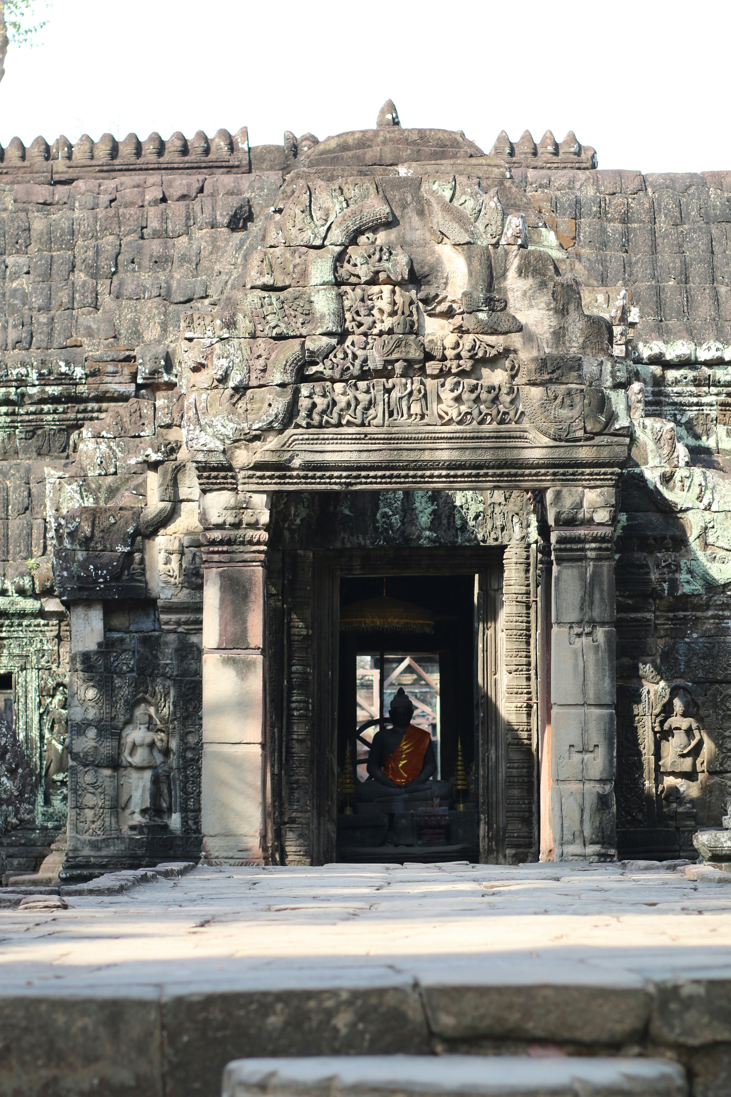 Intricate stone carvings frame a doorway leading to a serene figure within a historic temple. The play of light highlights the textures and details of the ancient architecture.