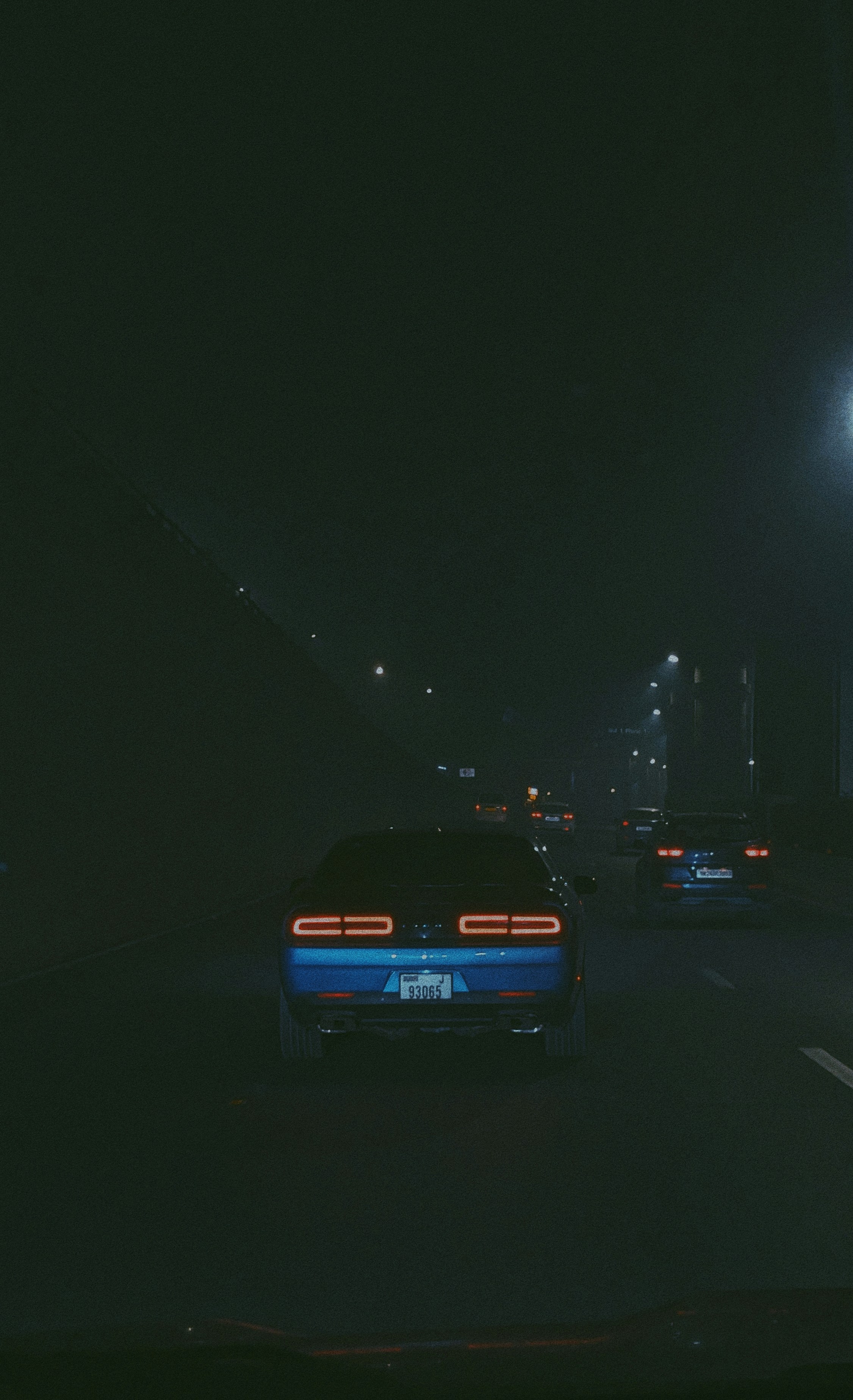 Nighttime street photograph of a blue muscle car in a tunnel, tail lights glowing as it follows other vehicles. Dim walls and distant headlights add depth to the moody scene.