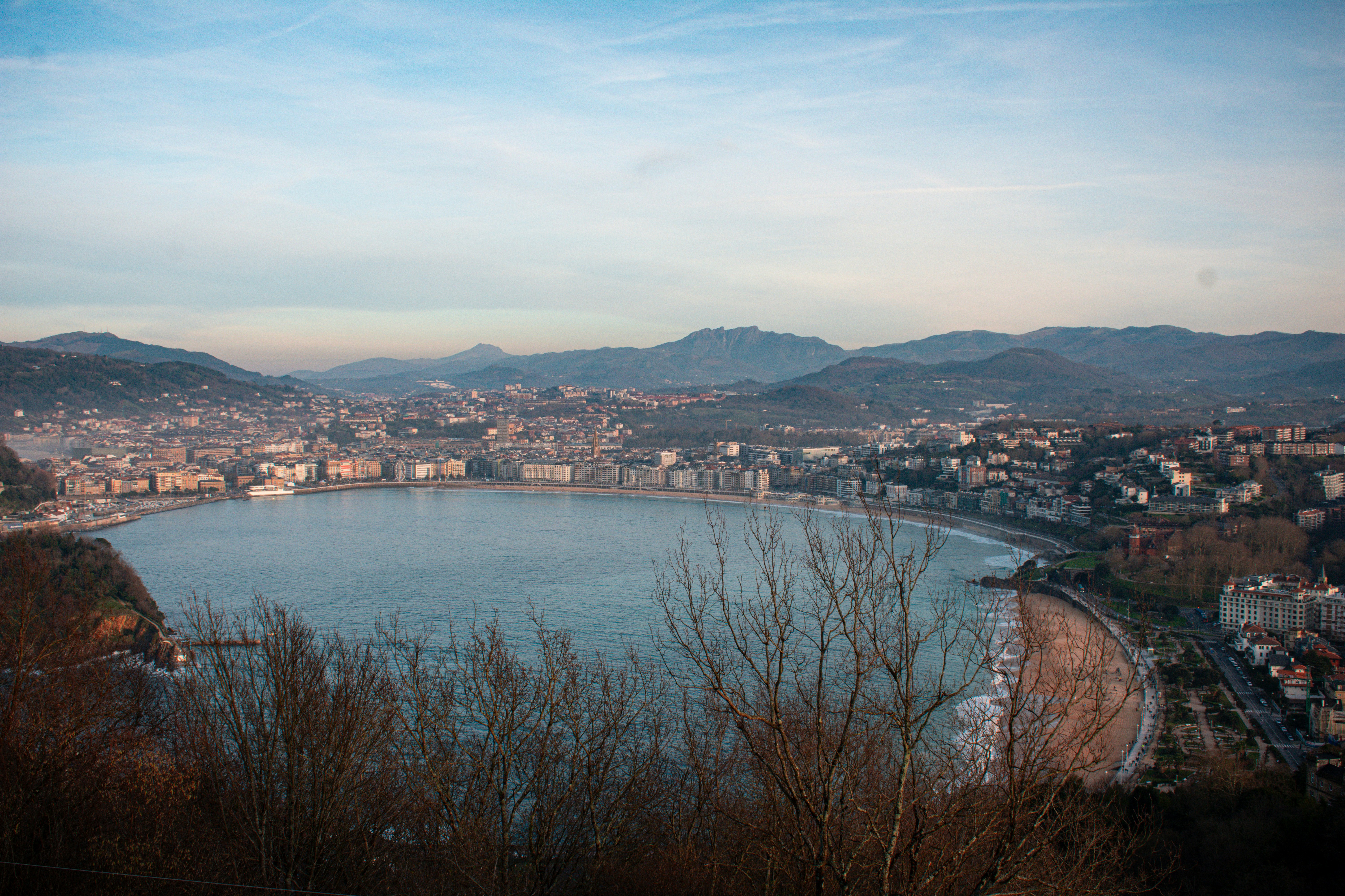 Curved bay bordered by urban landscape and distant mountains under a blue sky.