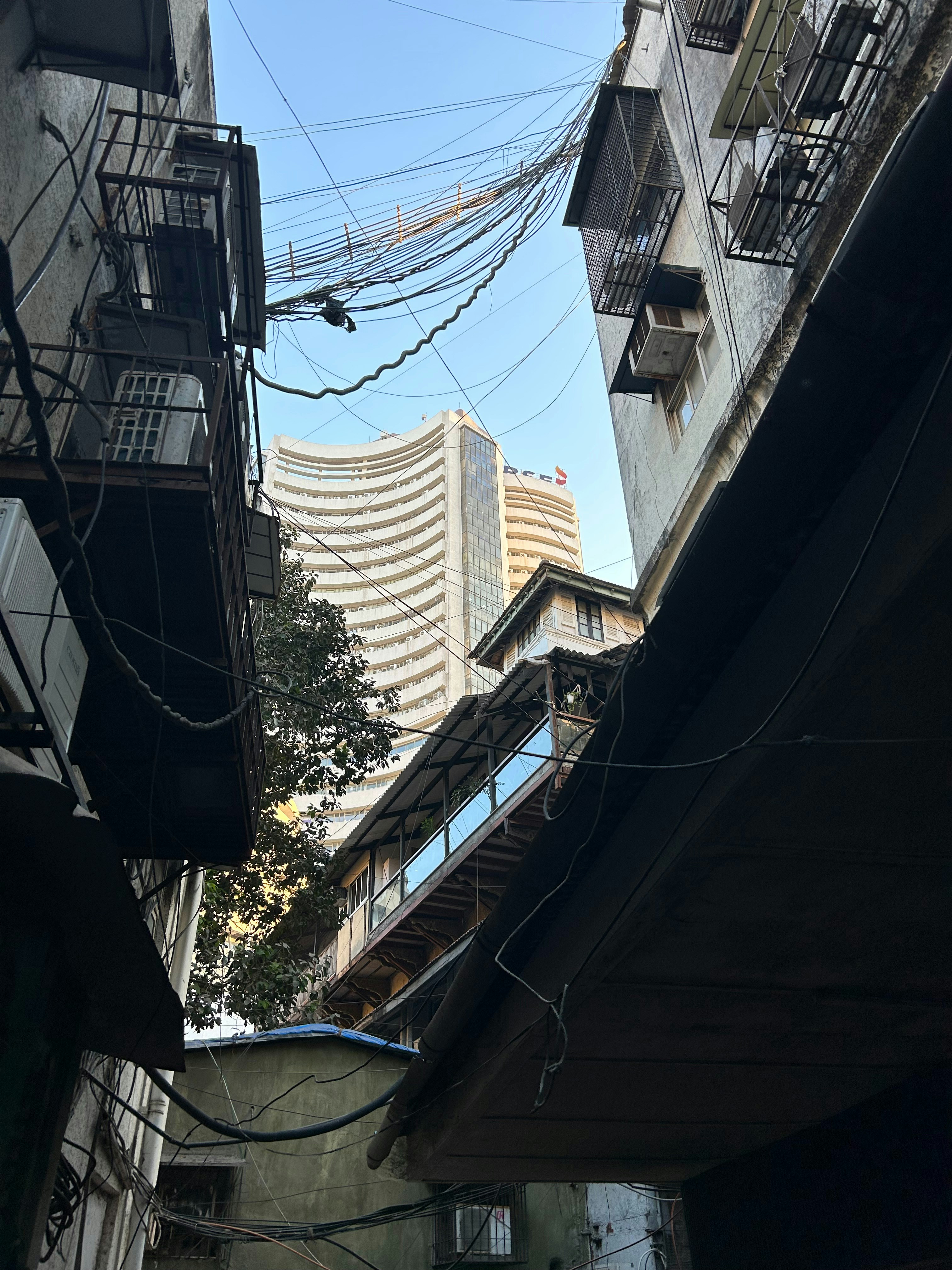 Photograph of a narrow urban alleyway with tangled power lines, guiding the eye toward a distant curved office tower under a clear blue sky.