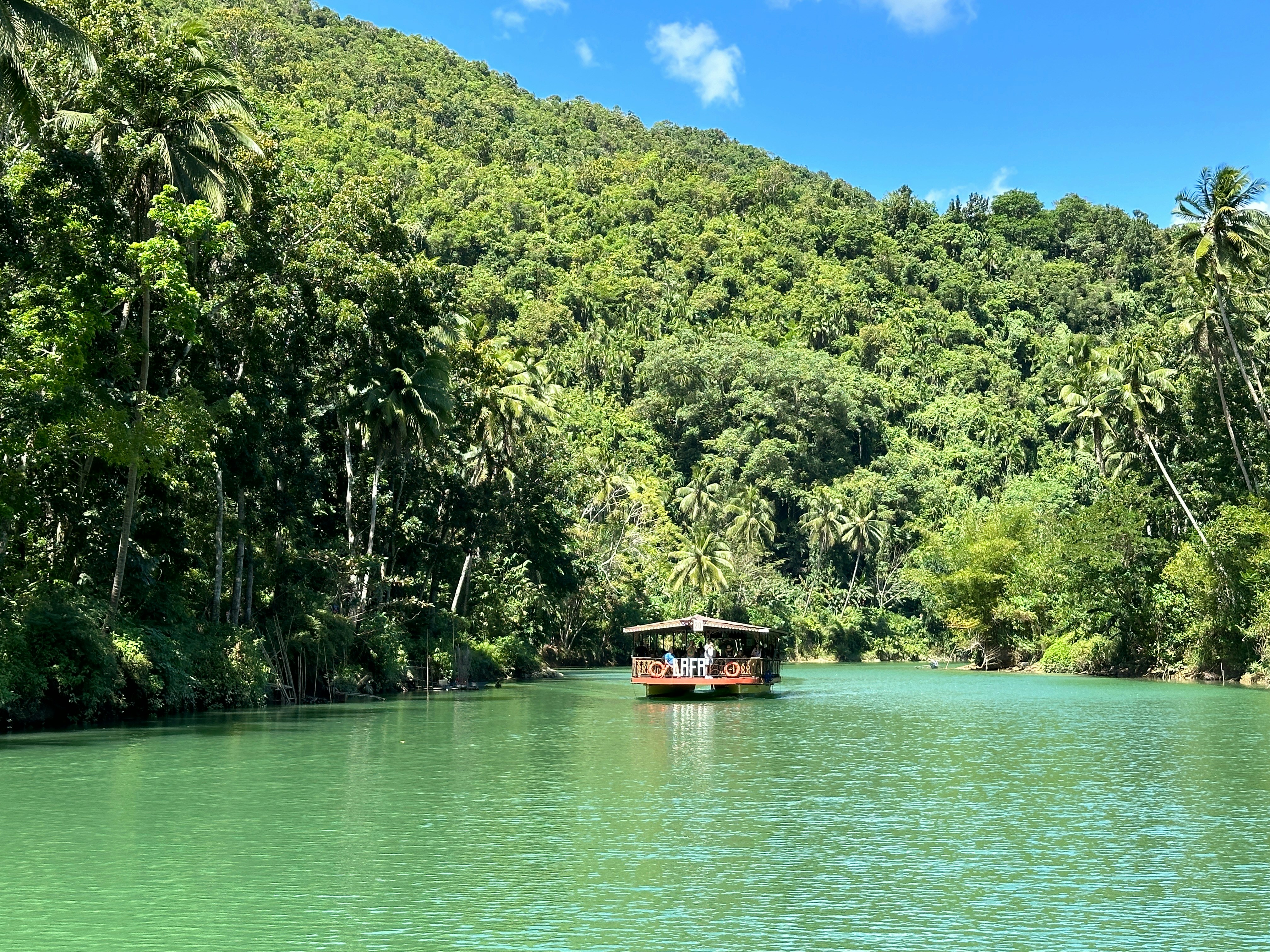 A traditional boat glides through the tranquil emerald waters, surrounded by lush tropical vegetation and rolling hills. The scene captures the essence of nature's peace.