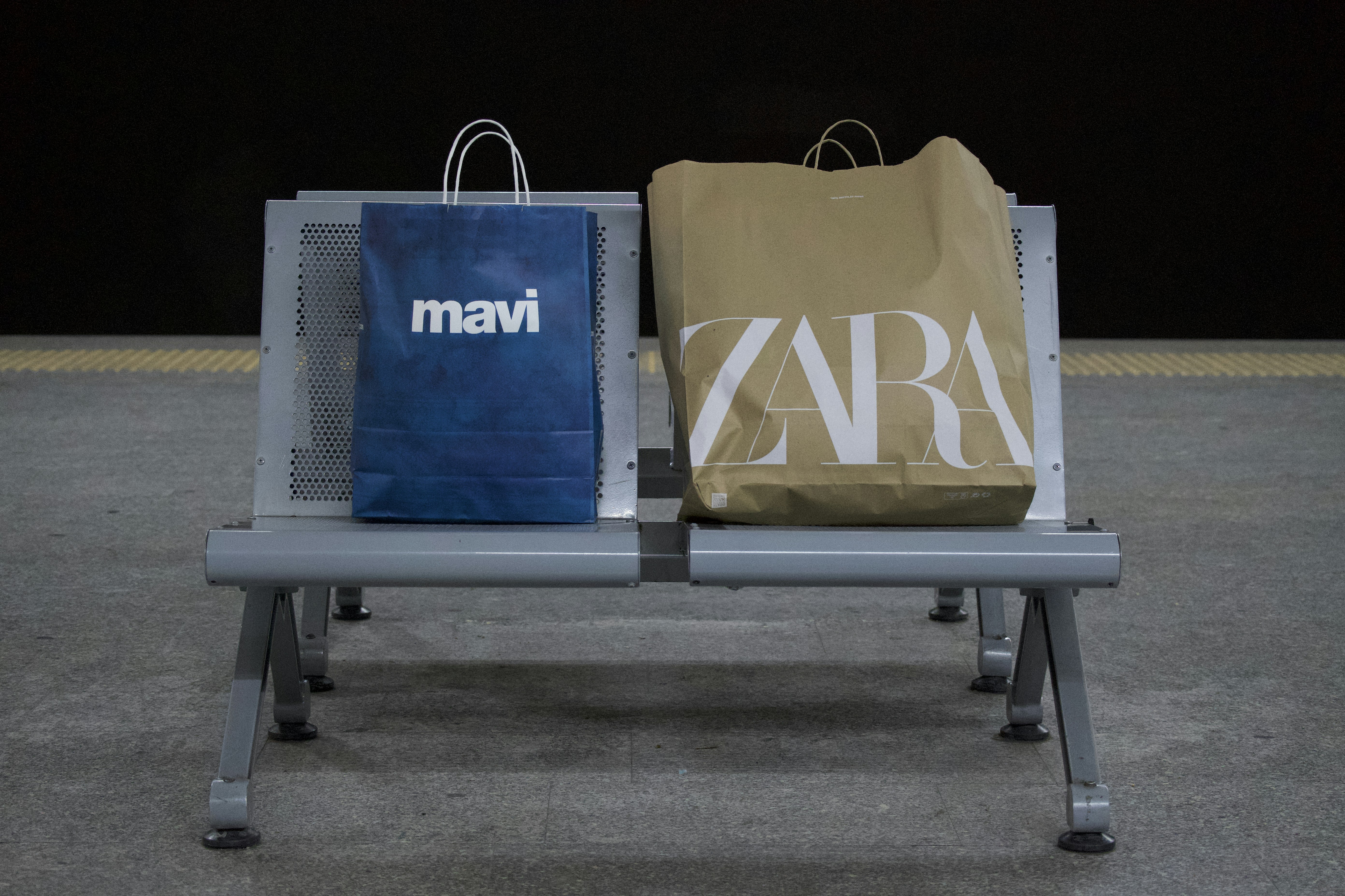 Two shopping bags, one blue from mavi and one beige from Zara, rest on a sleek bench in an urban setting.
