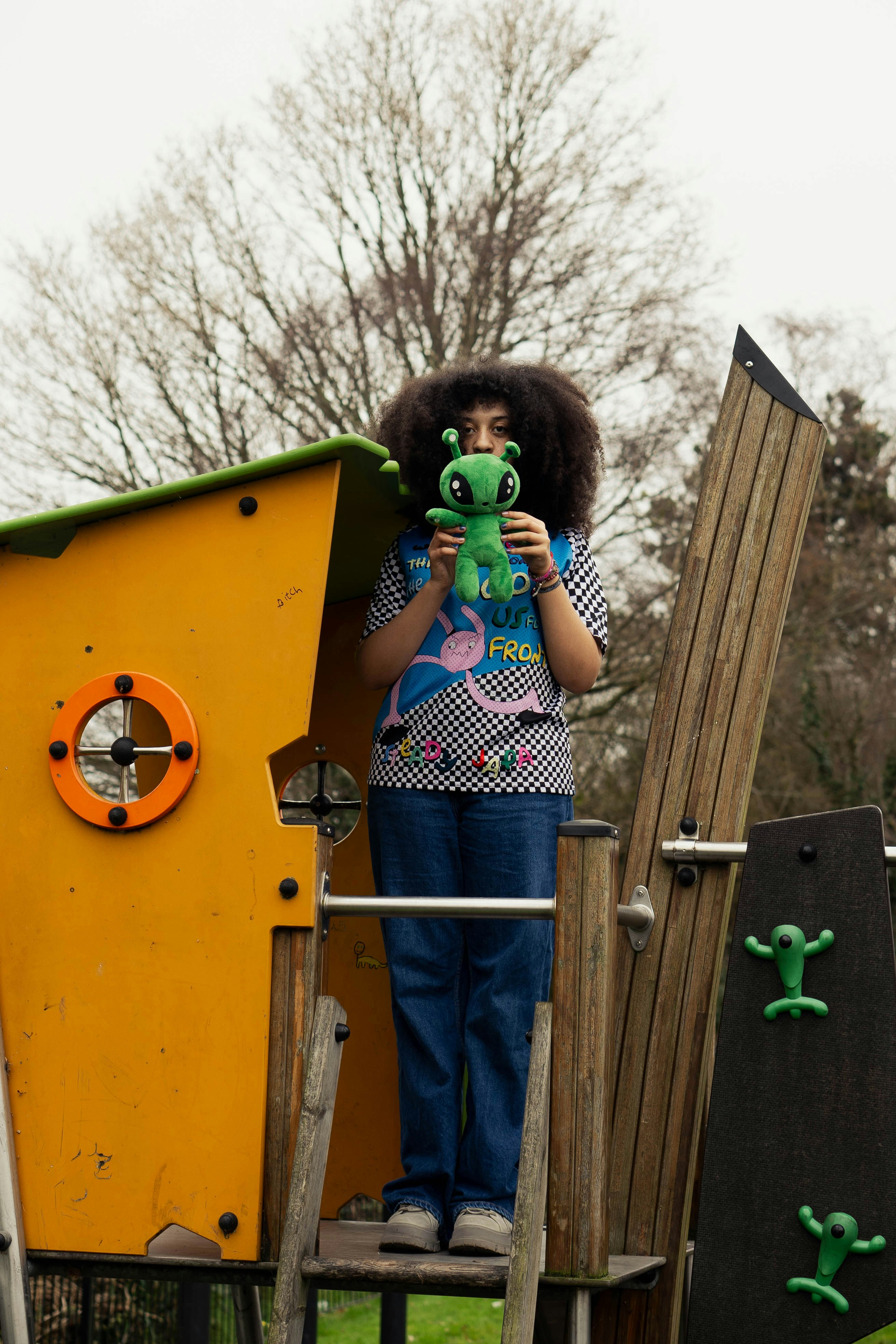 Child on playground platform holds a green plush toy amid colorful equipment