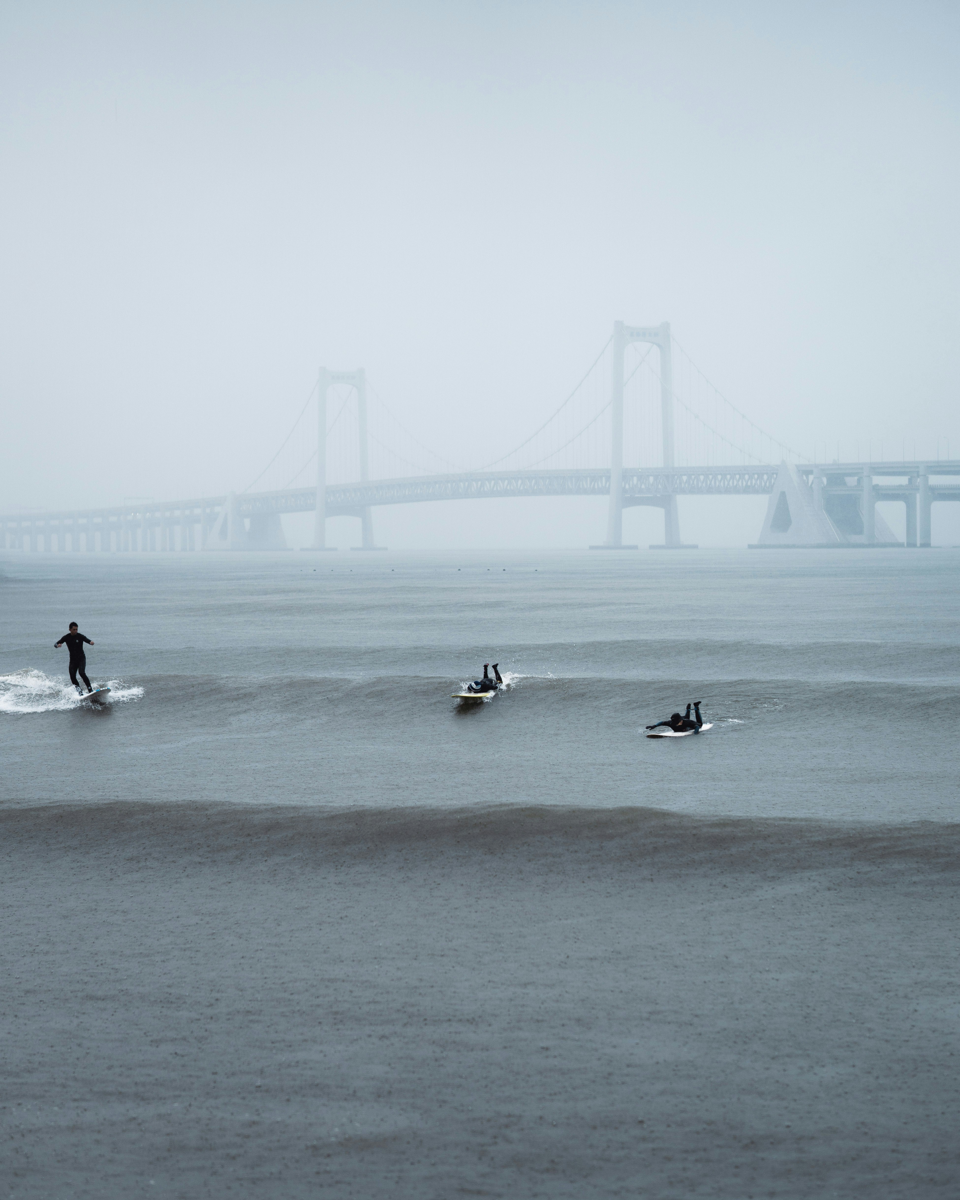 Fogbound Surfers: Three Ride the Quiet Atlantic as a Bridge Emerges in the Mist