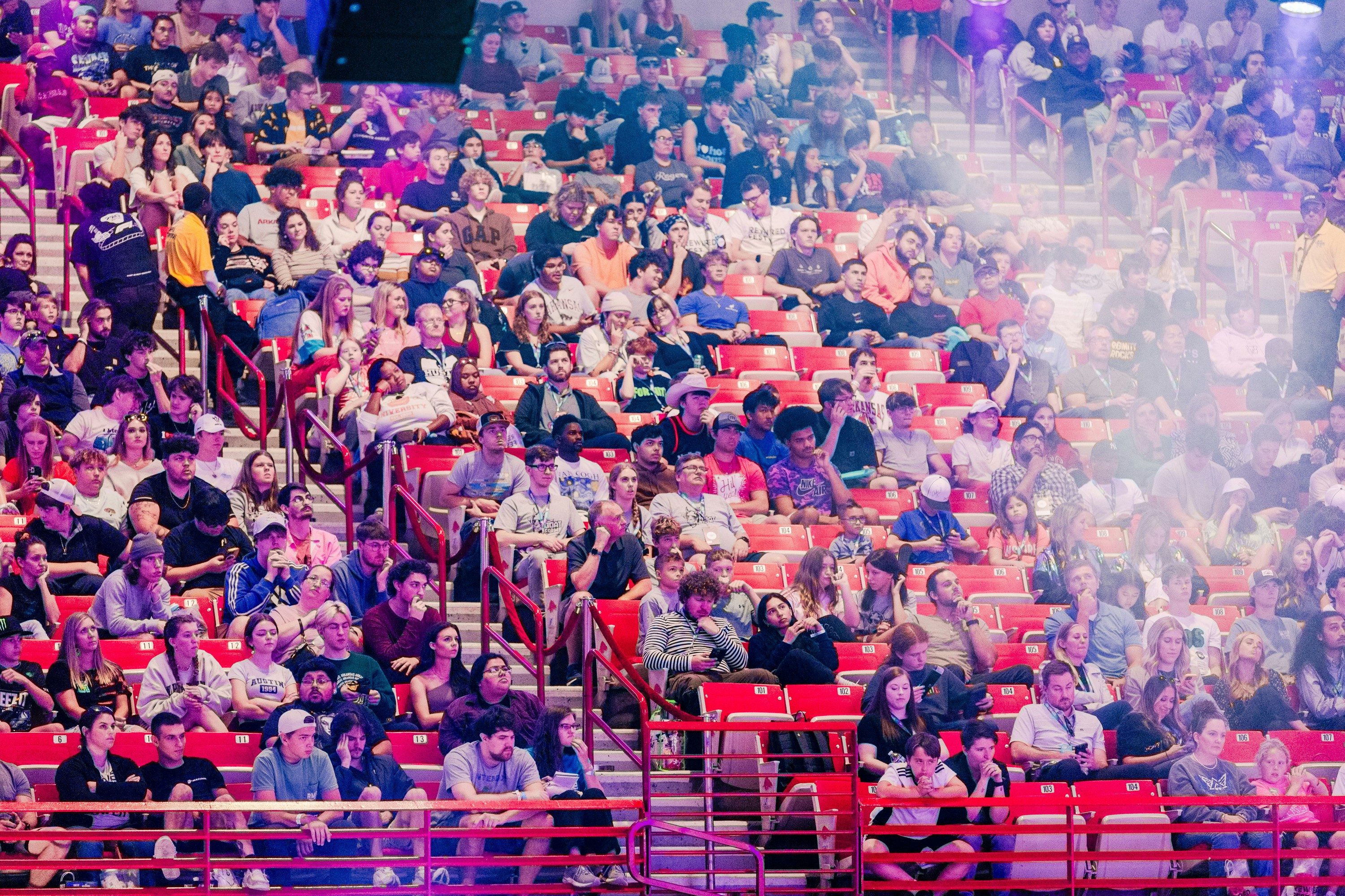 Large crowd seated in a stadium with colorful lights and haze creating a dynamic atmosphere.