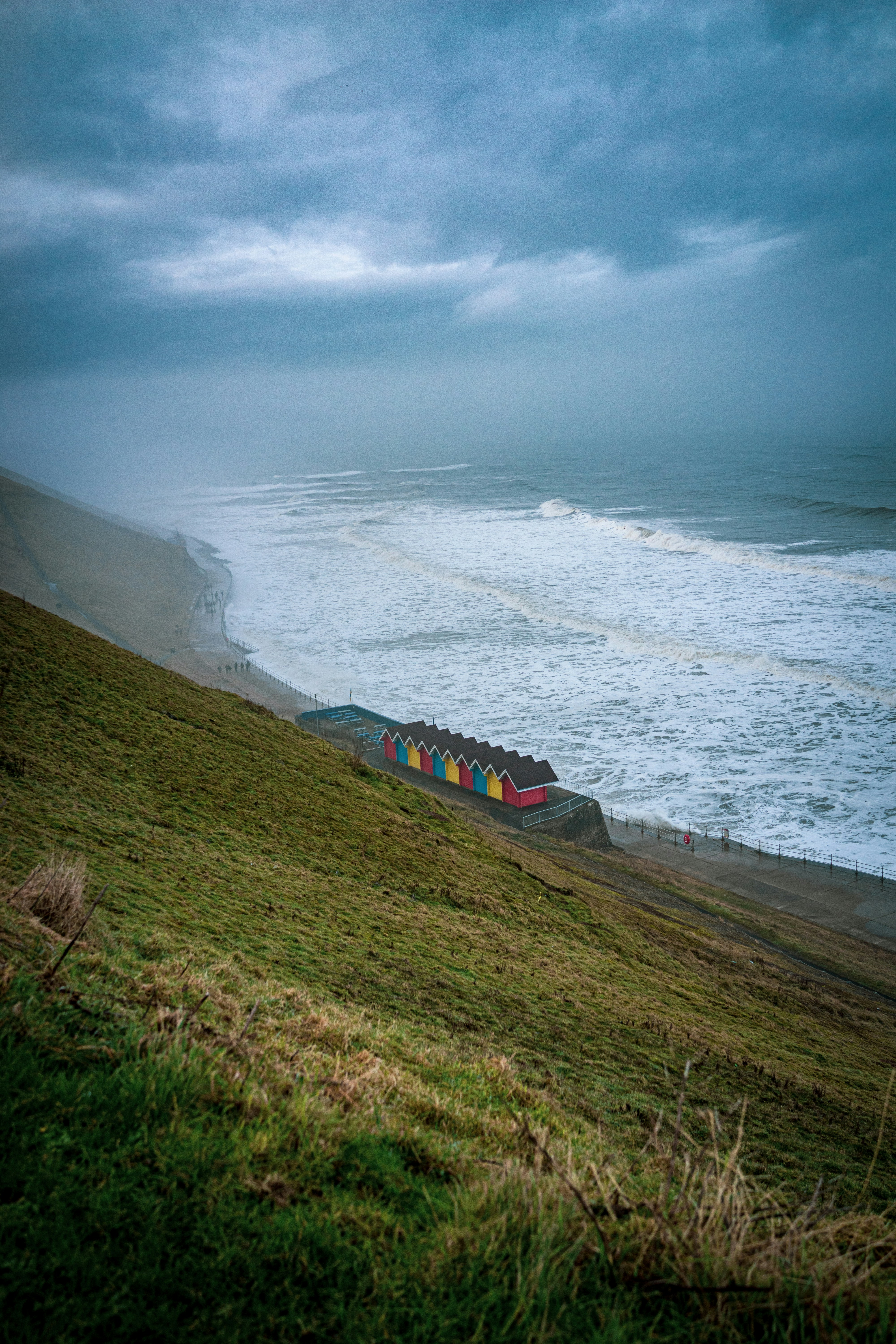 Colorful beach huts line a windswept cliff along a foaming shoreline under a moody sky.