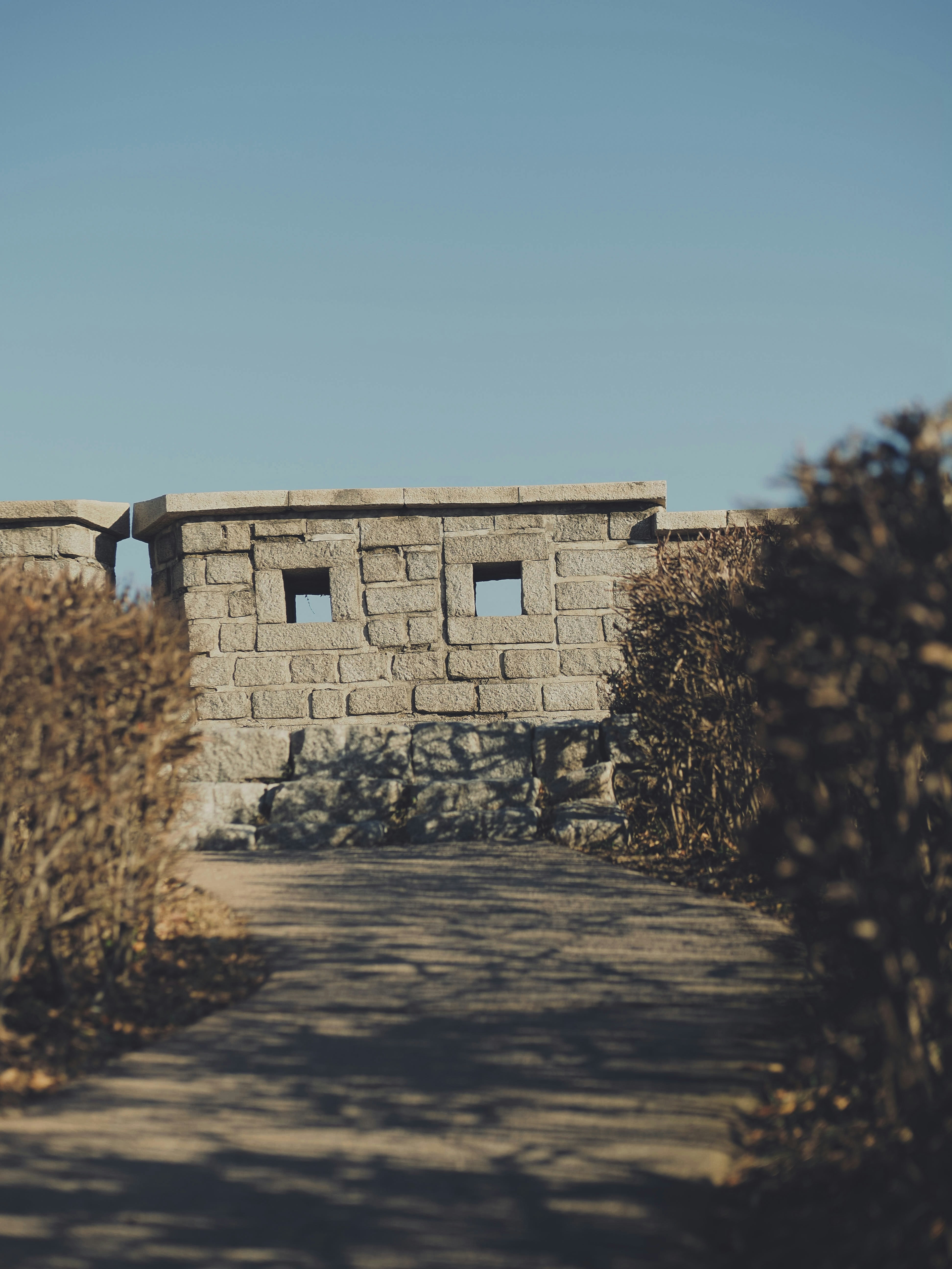 Sunlit gravel path leads to a weathered stone wall with two square openings, framed by clipped hedges and a clear blue sky.