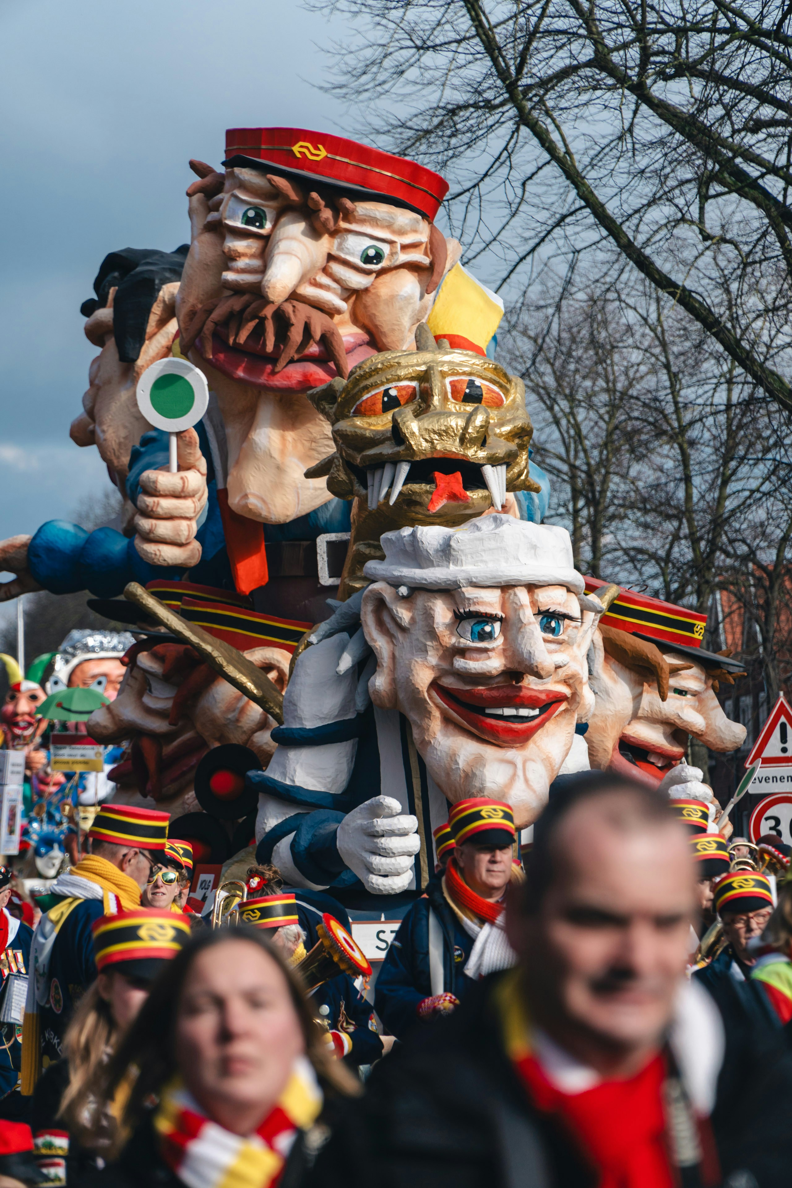 A colorful carnival parade with a large float depicting exaggerated, cartoon-like figures. One of the main figures is dressed as a train conductor, holding a green signal disc, with a mustache and an oversized nose. Another figure, positioned below, has a smiling face and is holding a golden lion's head. People in the crowd are wearing matching striped hats and scarves, and the festive atmosphere is lively and cheerful. The parade takes place outdoors on a partly cloudy day, with trees visible in the background.