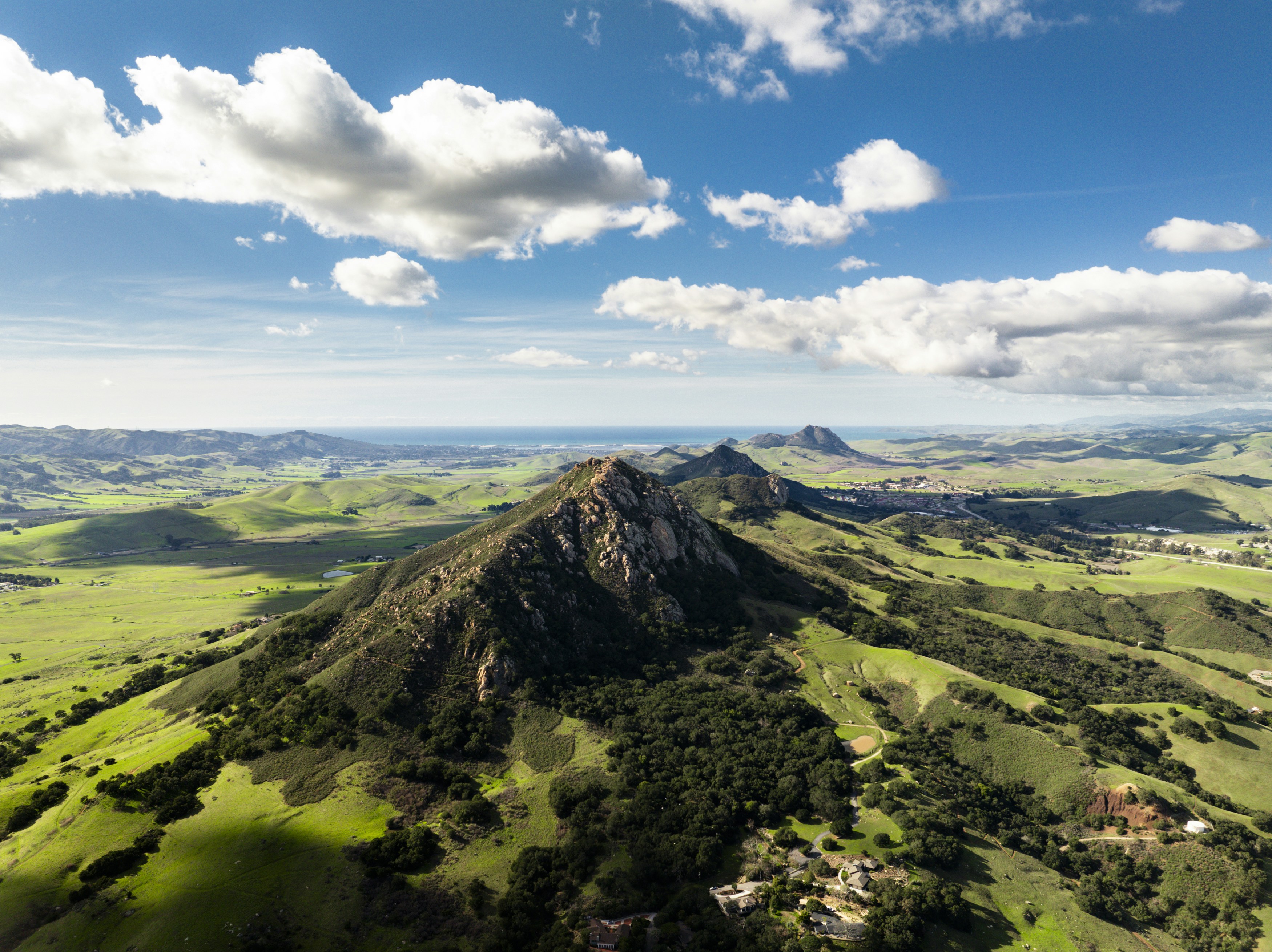 Aerial view of lush green hills and a rugged mountain range beneath a sky dotted with fluffy clouds.