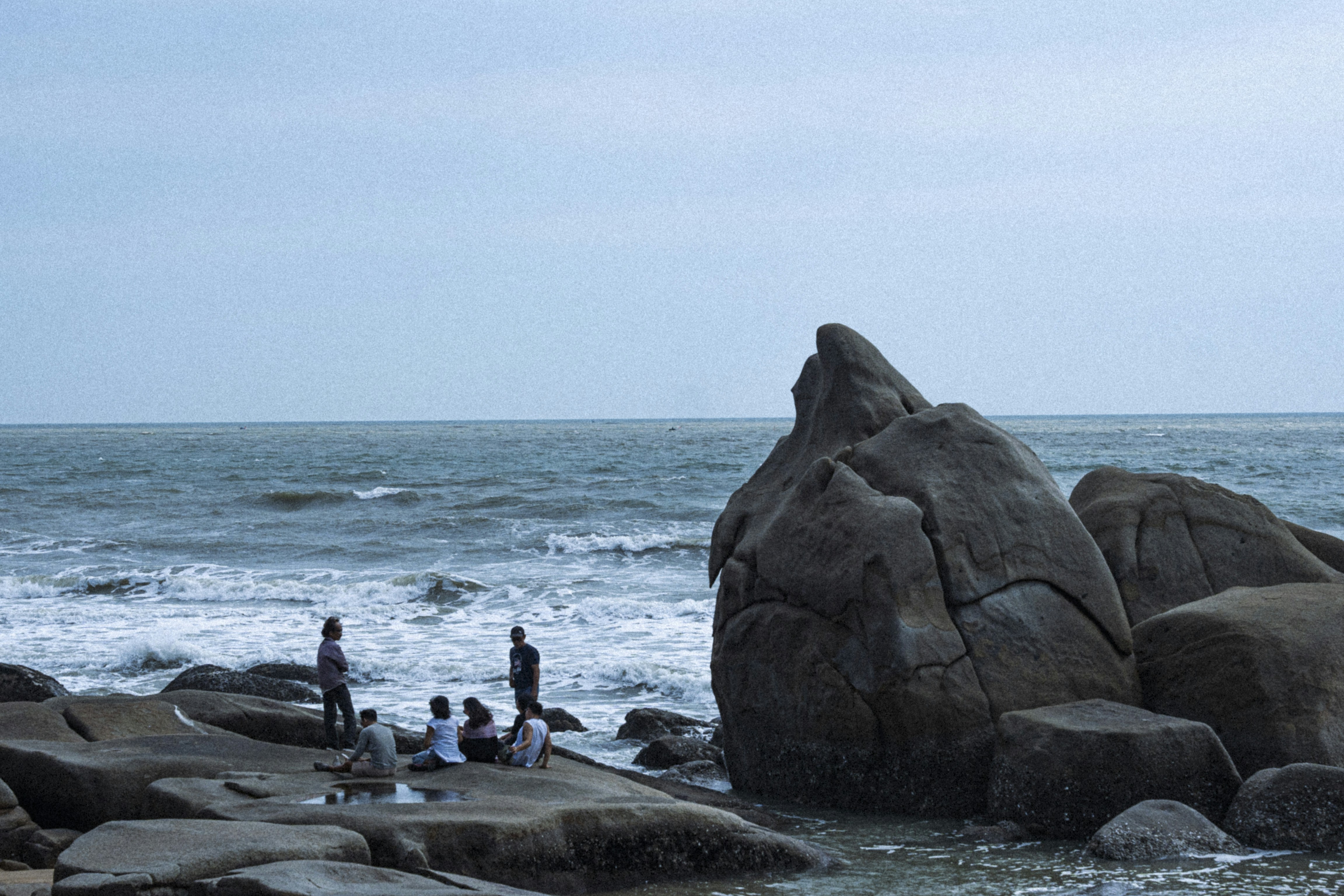 Group of people standing on large coastal rocks by the ocean under a cloudy sky.