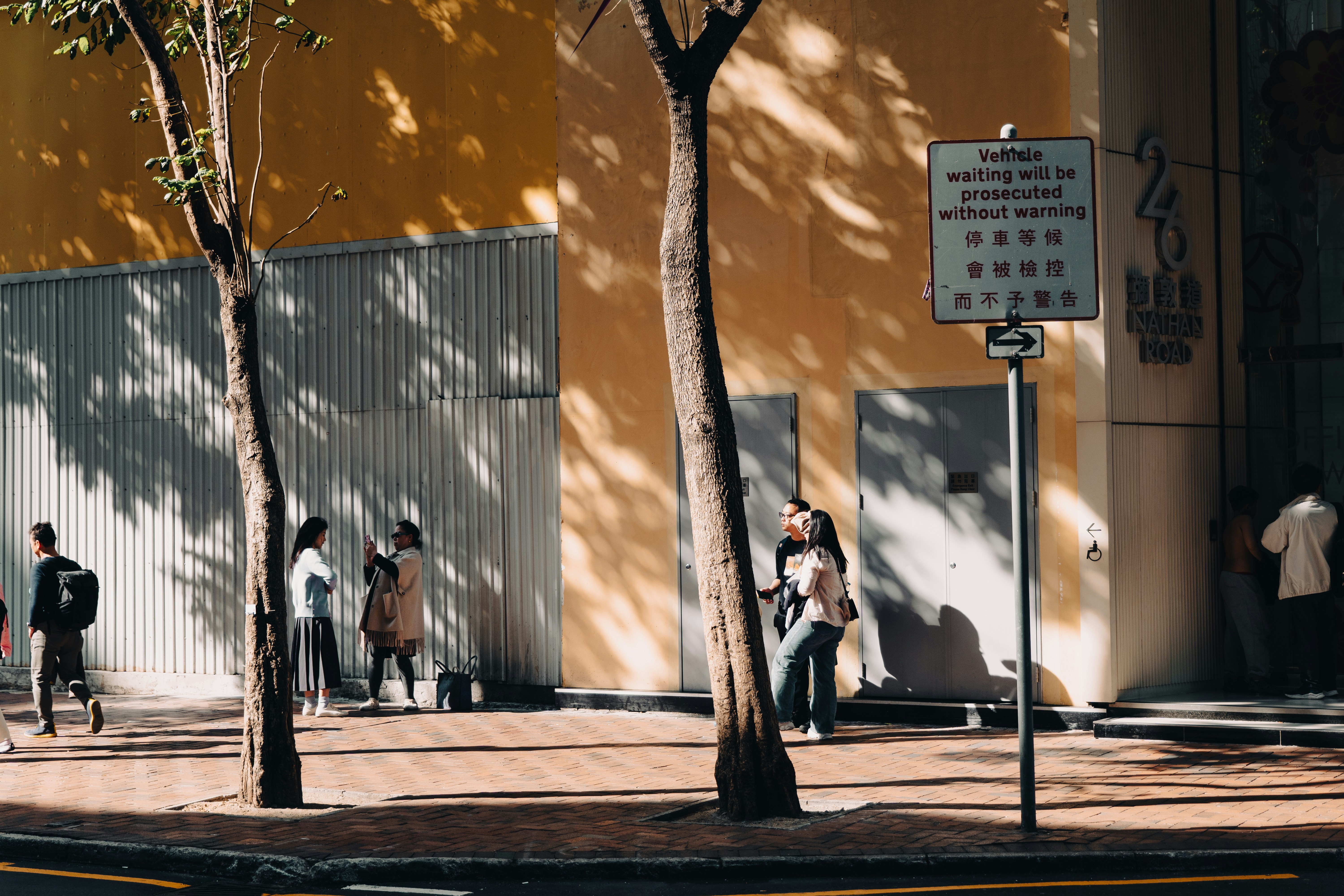 People walking along a sunlit urban street, with shadows cast by trees and buildings creating a dynamic interplay of light and form.