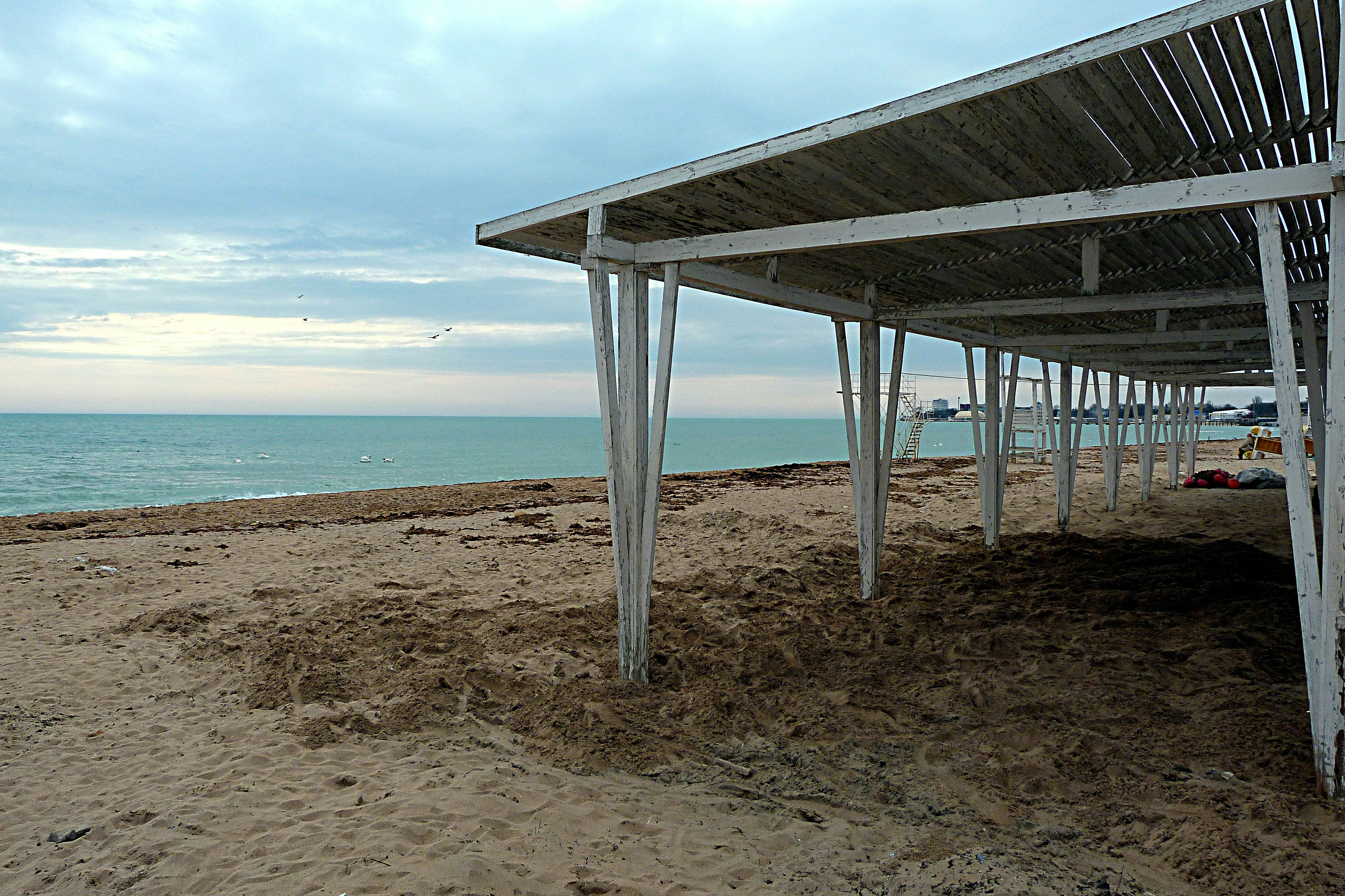 A photograph showing weathered wooden posts forming a shelter corridor along a sandy beach, guiding the eye toward the calm sea.