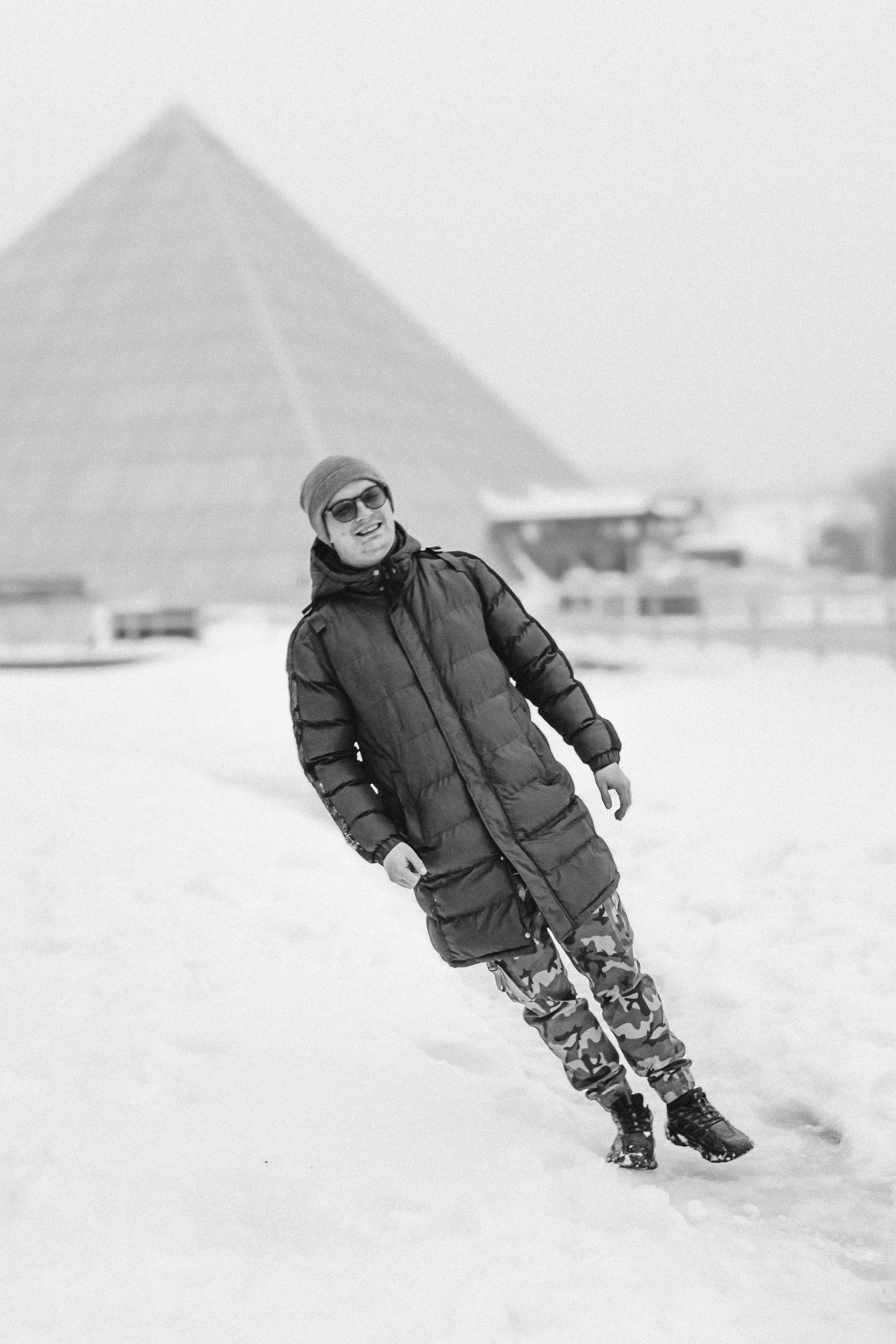 A person dressed in winter attire stands playfully in the snow, with a striking pyramid structure in the background.
