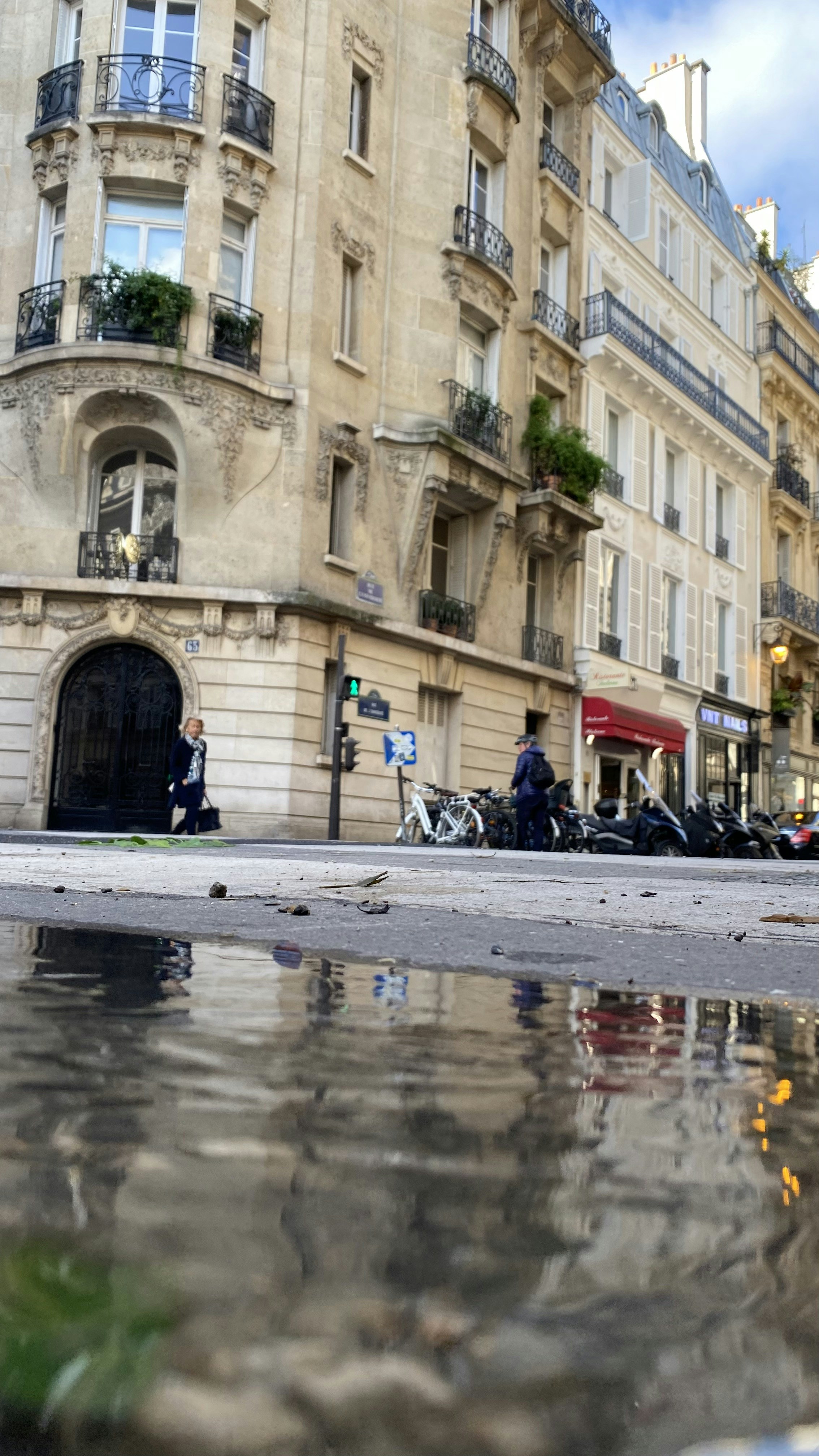 A street scene capturing a puddle of water reflecting the surroundings and the passing pedestrians and vehicles.