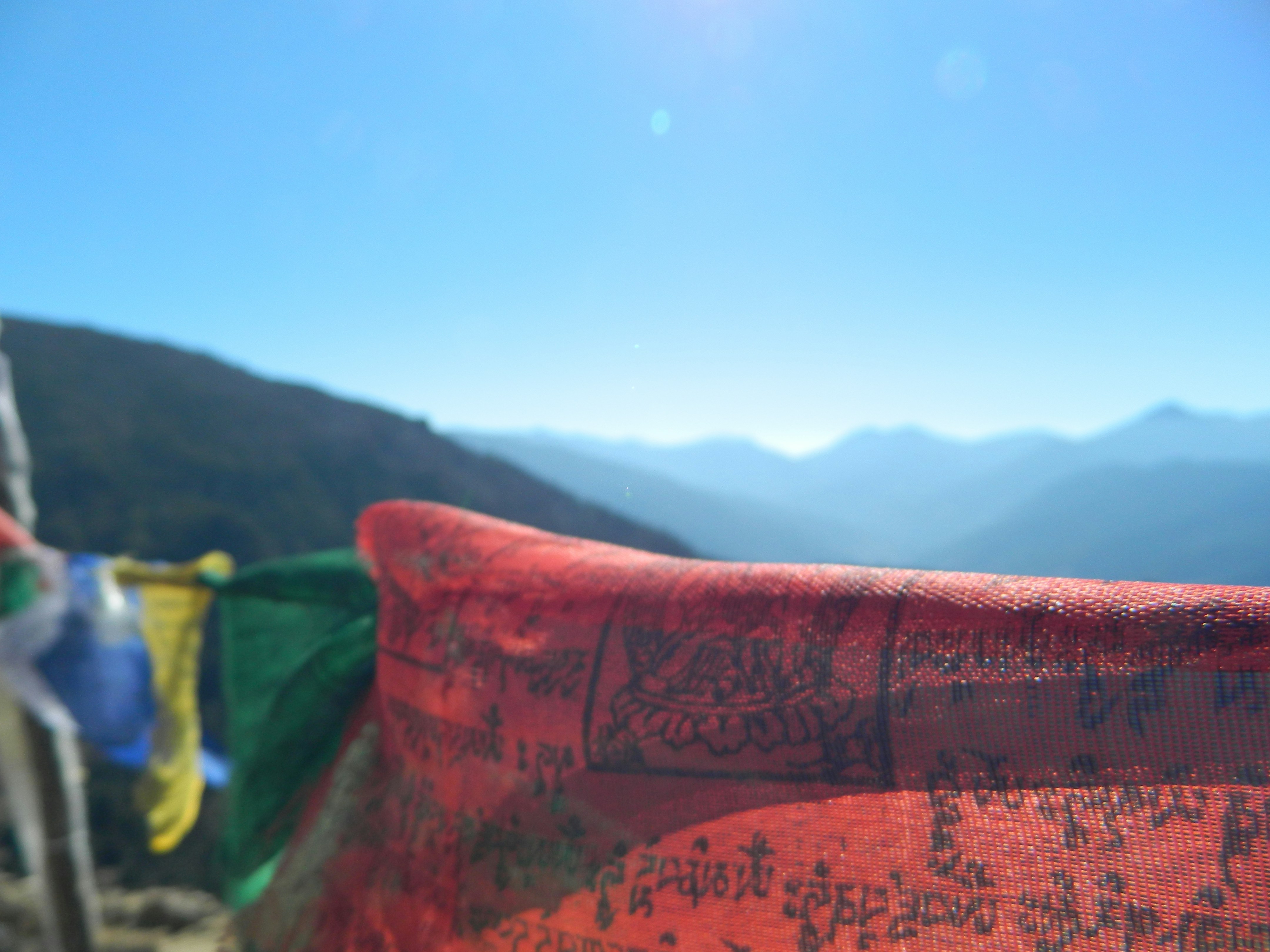 Colorful prayer flags flutter against a backdrop of misty mountain ranges under a clear blue sky.