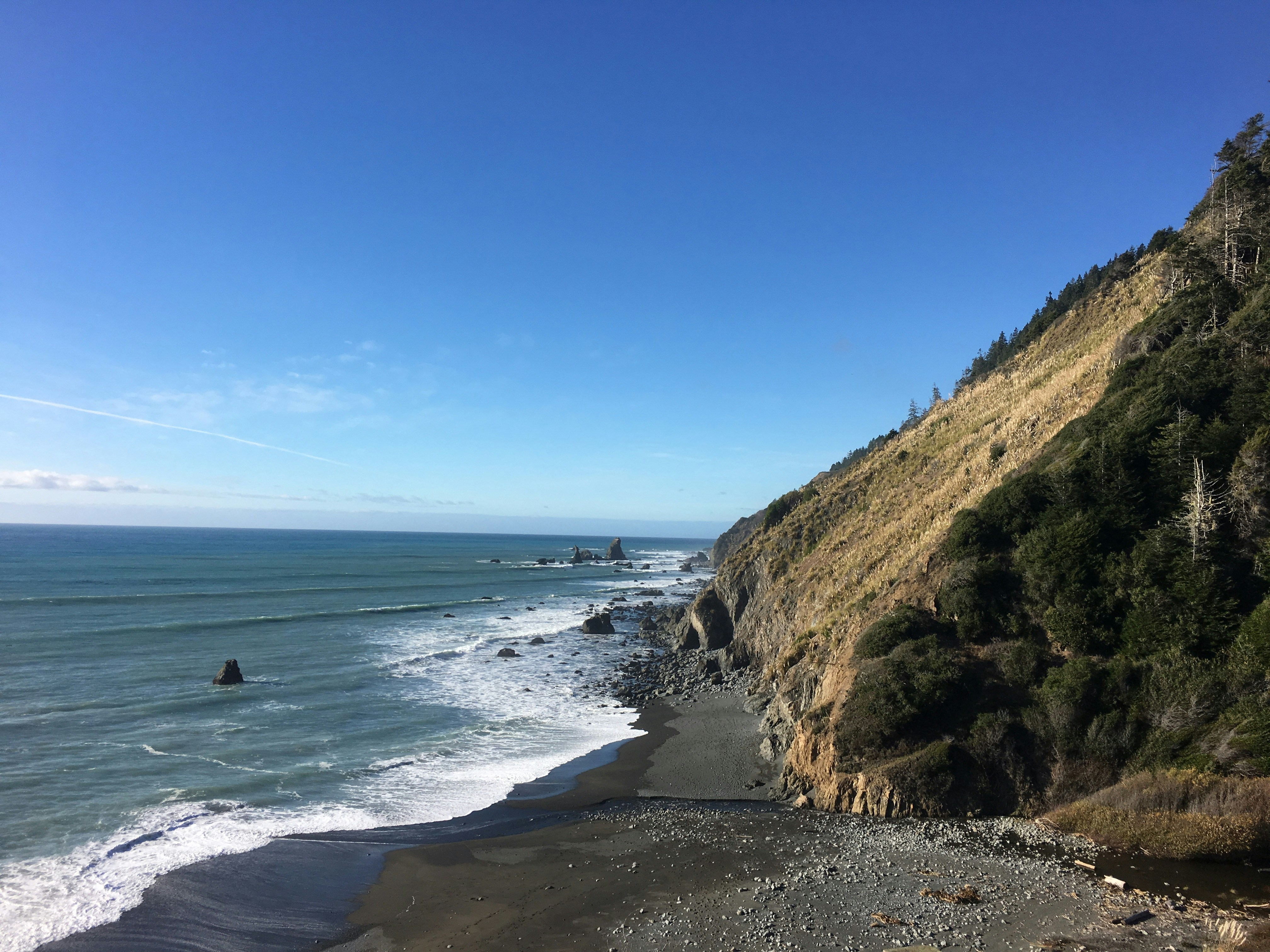 Expansive view of a rugged coastline with waves gently crashing on the shore, framed by a lush hillside and clear blue sky.
