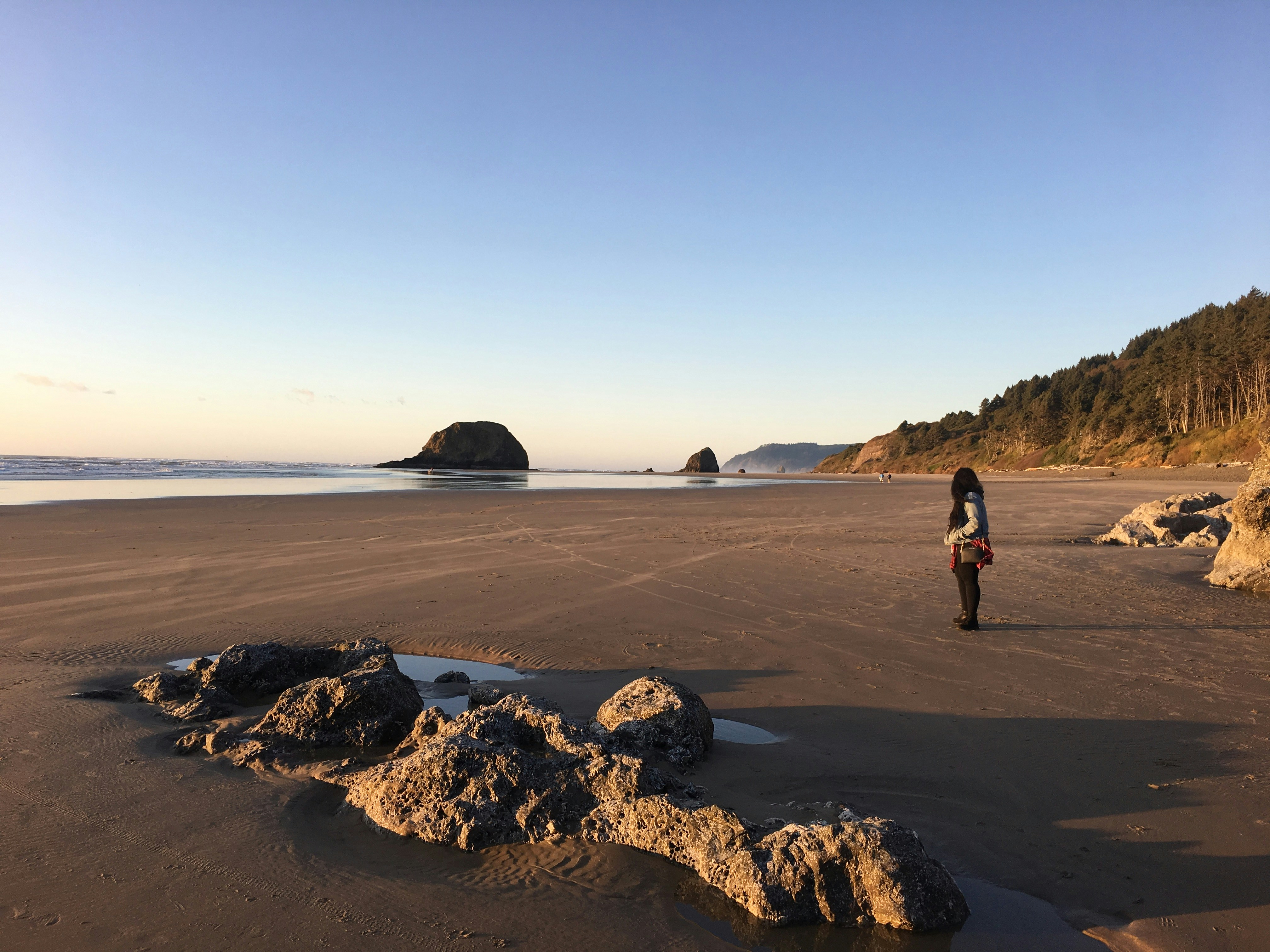 Person standing on Arcadia Beach during sunset with sea stacks and distant forested cliffs.