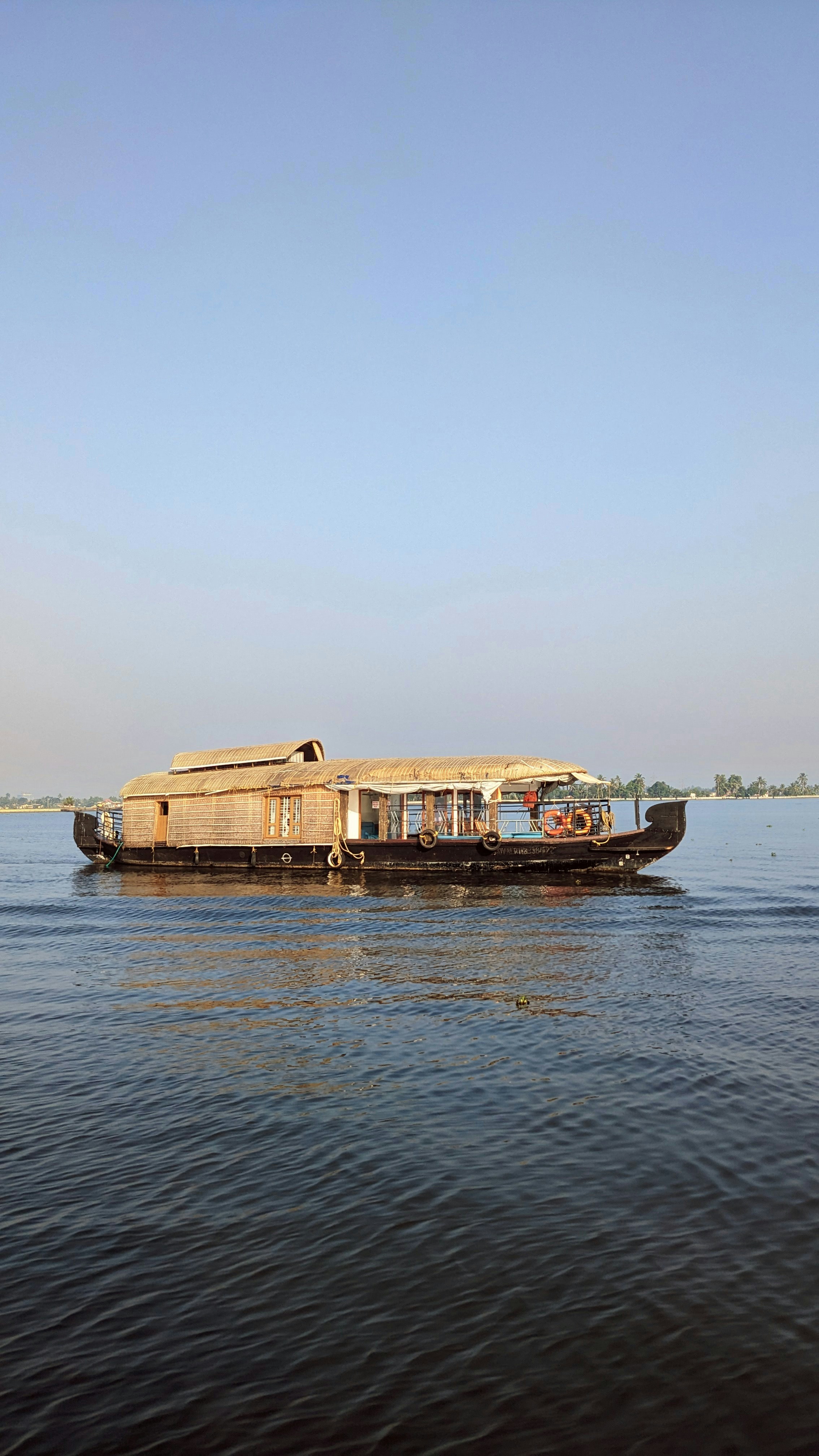 A wooden houseboat boat with a thatched roof floats serenely on a calm body of backwaters in Alleppey Kerala, surrounded by lush greenery and a clear blue sky. | a long boat is floating on the water