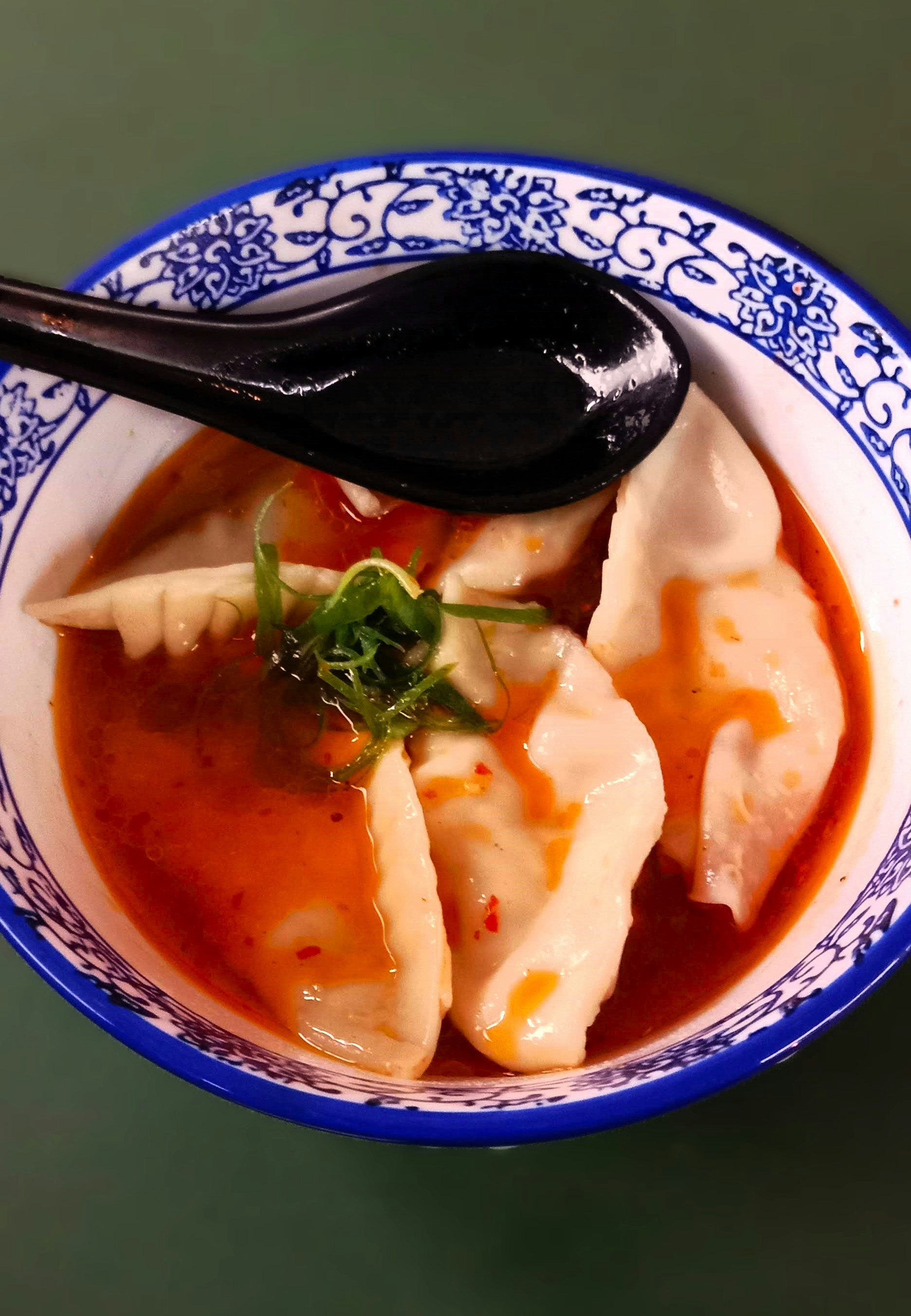 Delicately folded dumplings float in a bright red broth inside a blue-and-white patterned bowl, with a glossy black spoon resting across the rim. The shot highlights warmth, texture, and the bowl's distinctive pattern.