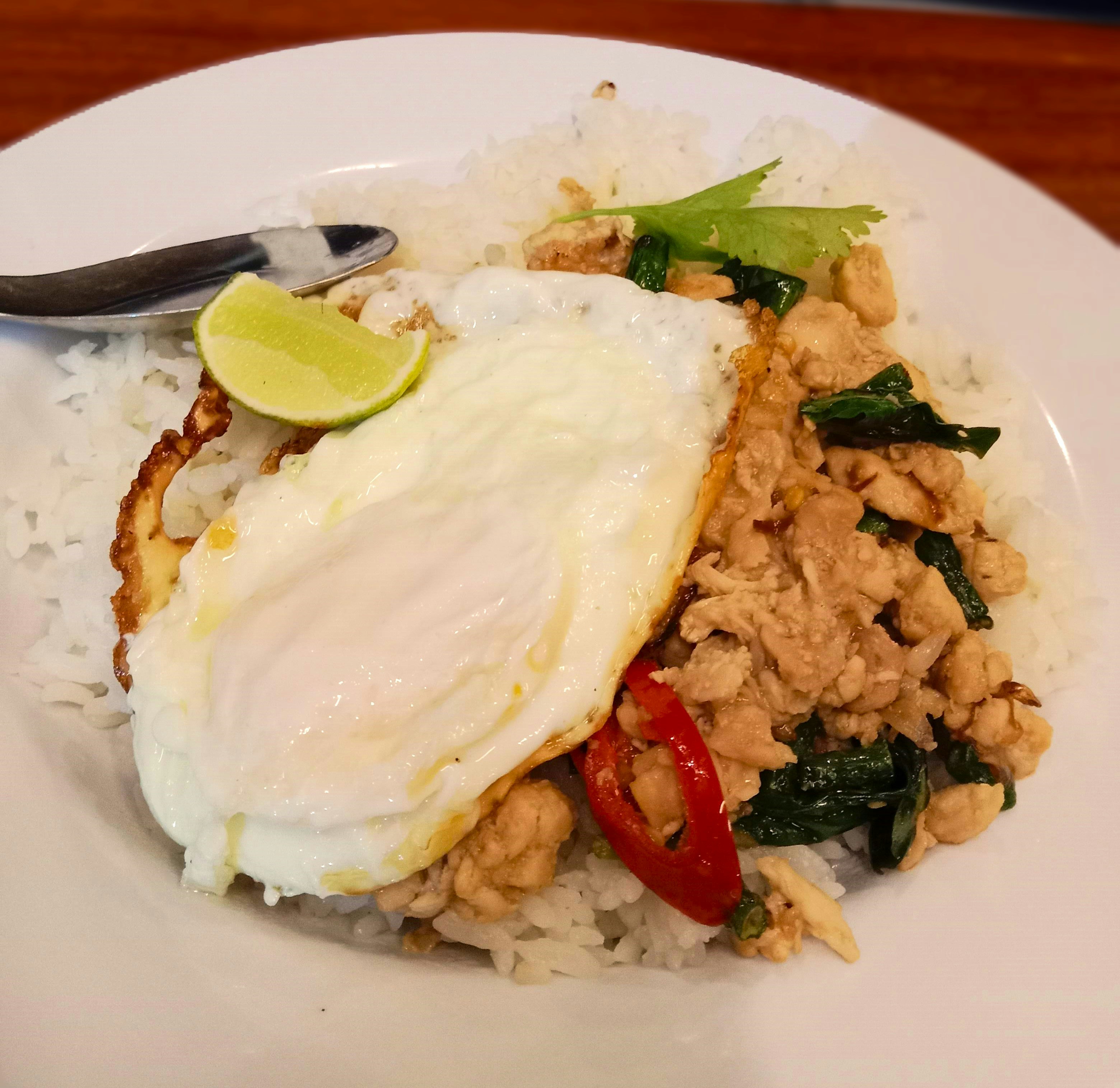 White plate of Thai basil chicken with rice, topped with a fried egg and garnished with a lime wedge.