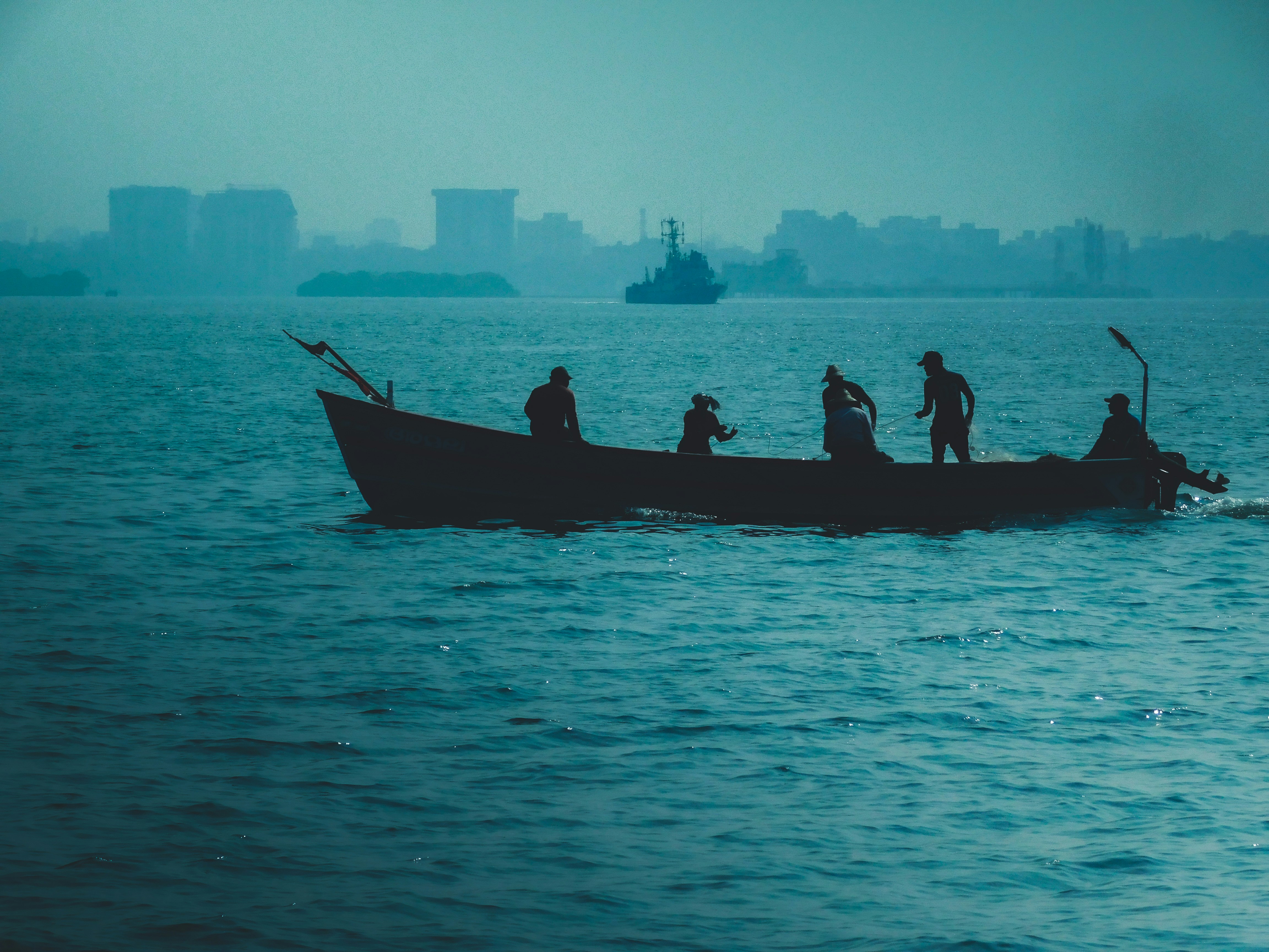 Silhouetted rowboat with several passengers glides across blue-hued water toward a distant city skyline. This photograph emphasizes stark contrasts and tranquil maritime mood.
