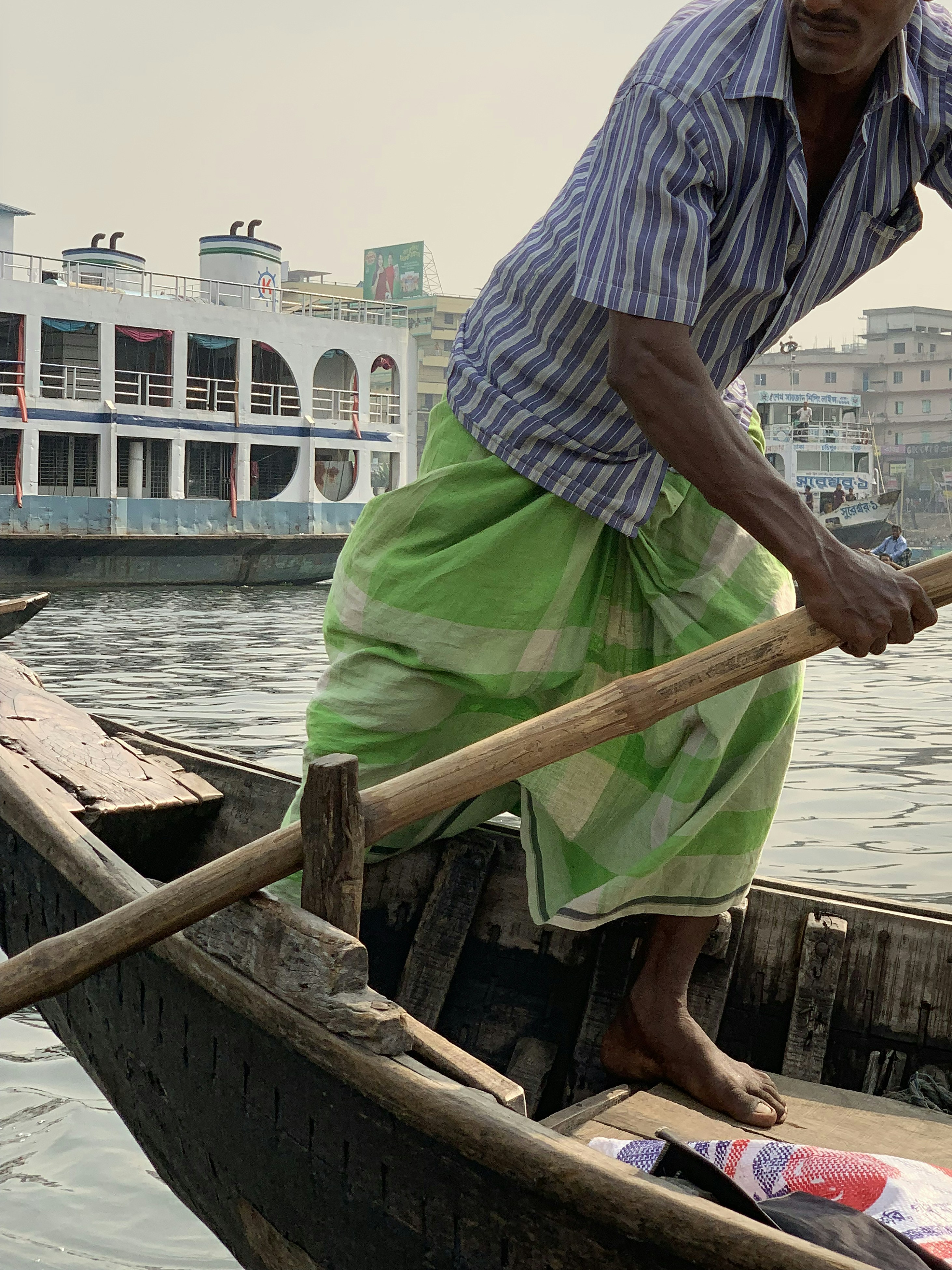 Rower in a Green Wrap Navigates a Weathered Boat Along a Busy Riverside