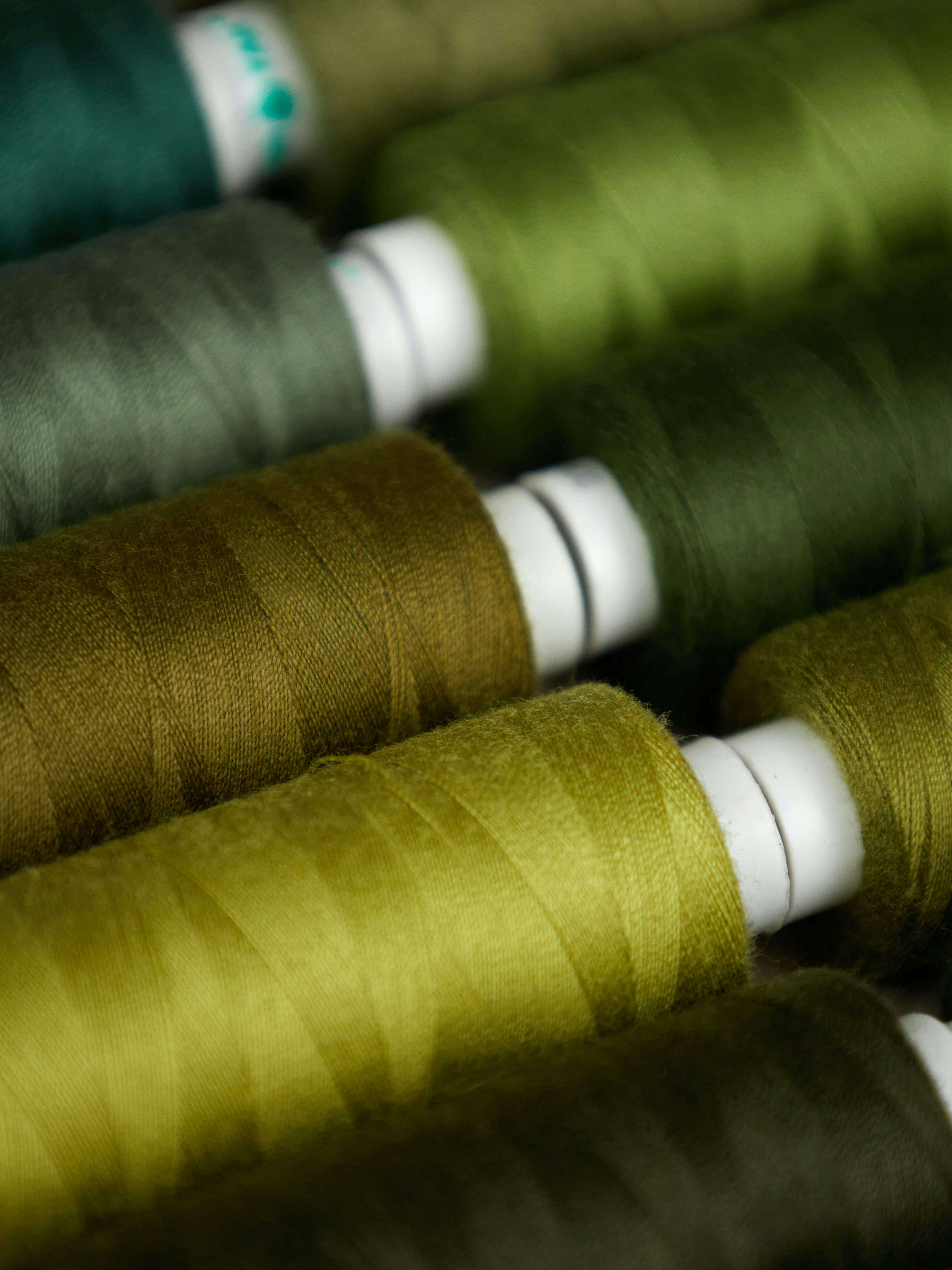 Spools of green sewing thread arranged in a gradient, showcasing various shades from deep forest to vibrant lime. The texture and color variation highlight the artistry of textile design.