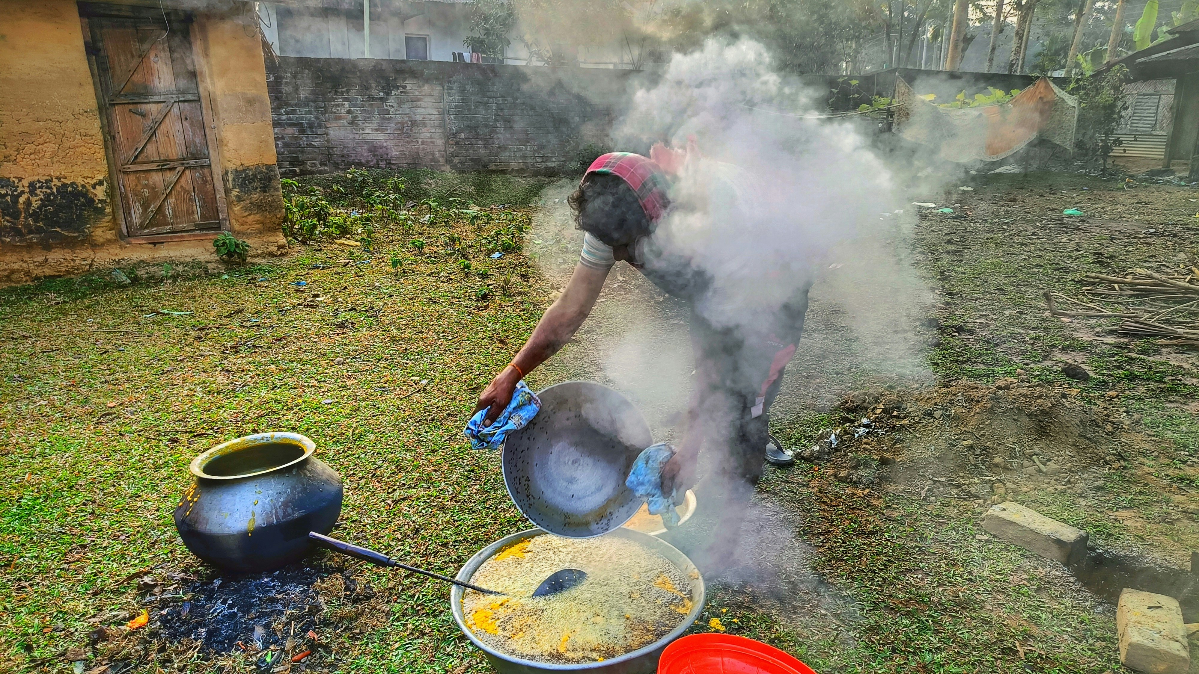 A person tends bubbling pots in an open-air cooking setup, steam rising in a dusty rural yard.