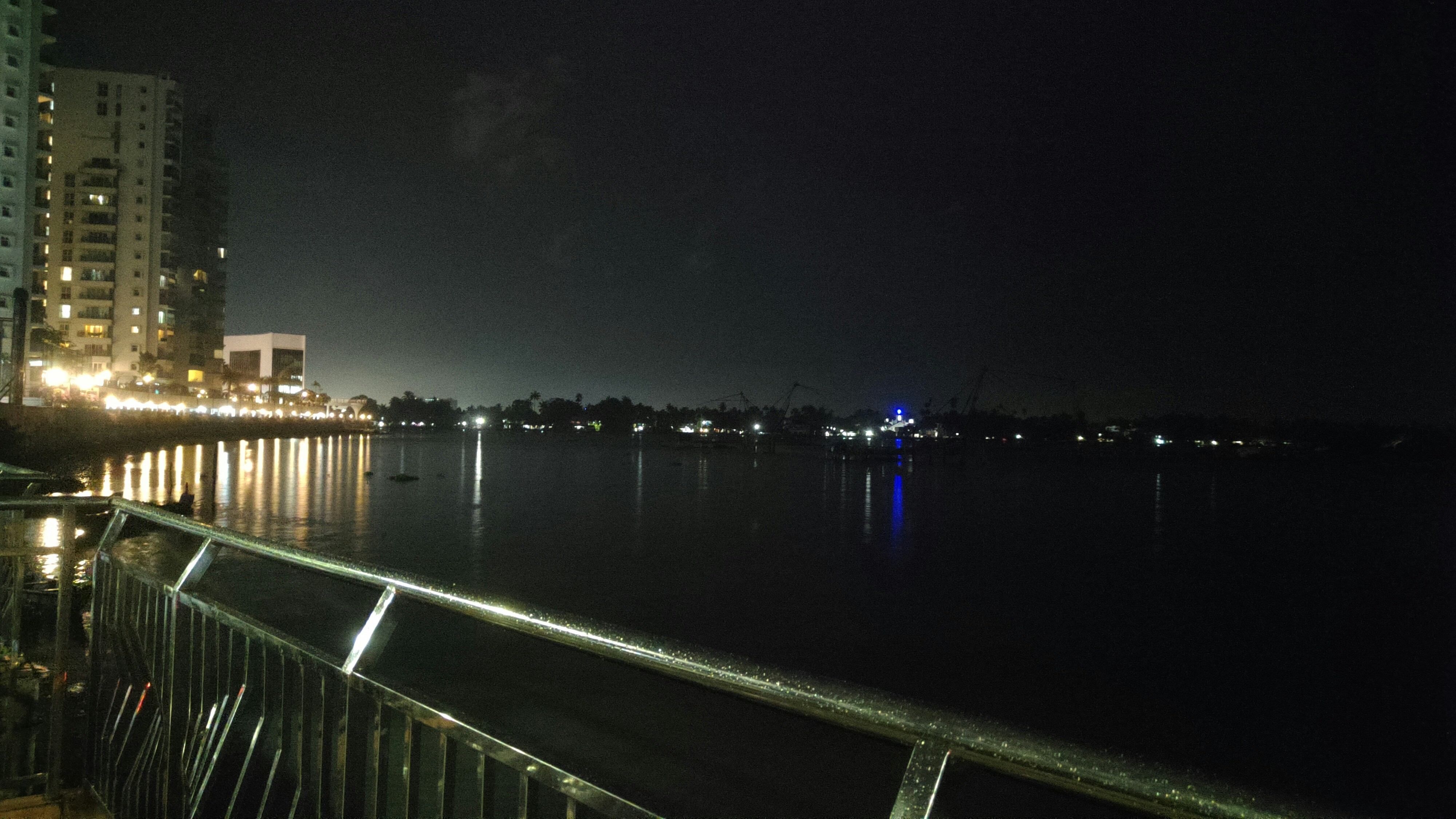 Night photograph of a waterfront cityscape with a metal railing in the foreground and reflections along the water. The composition emphasizes the distant skyline lit by street and building lights.