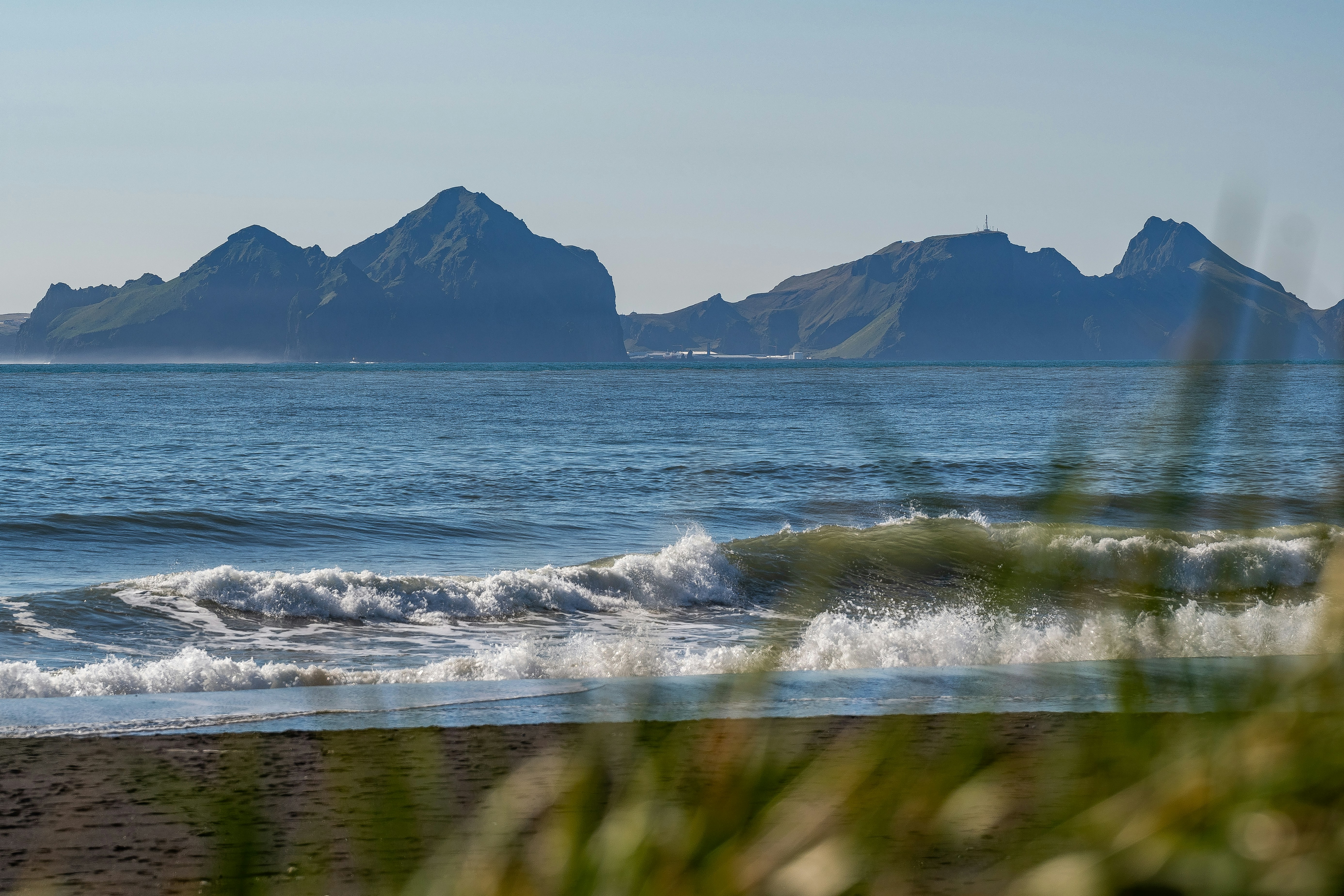 Ocean waves gently crash against a sandy shore with distant volcanic islands under a clear sky.