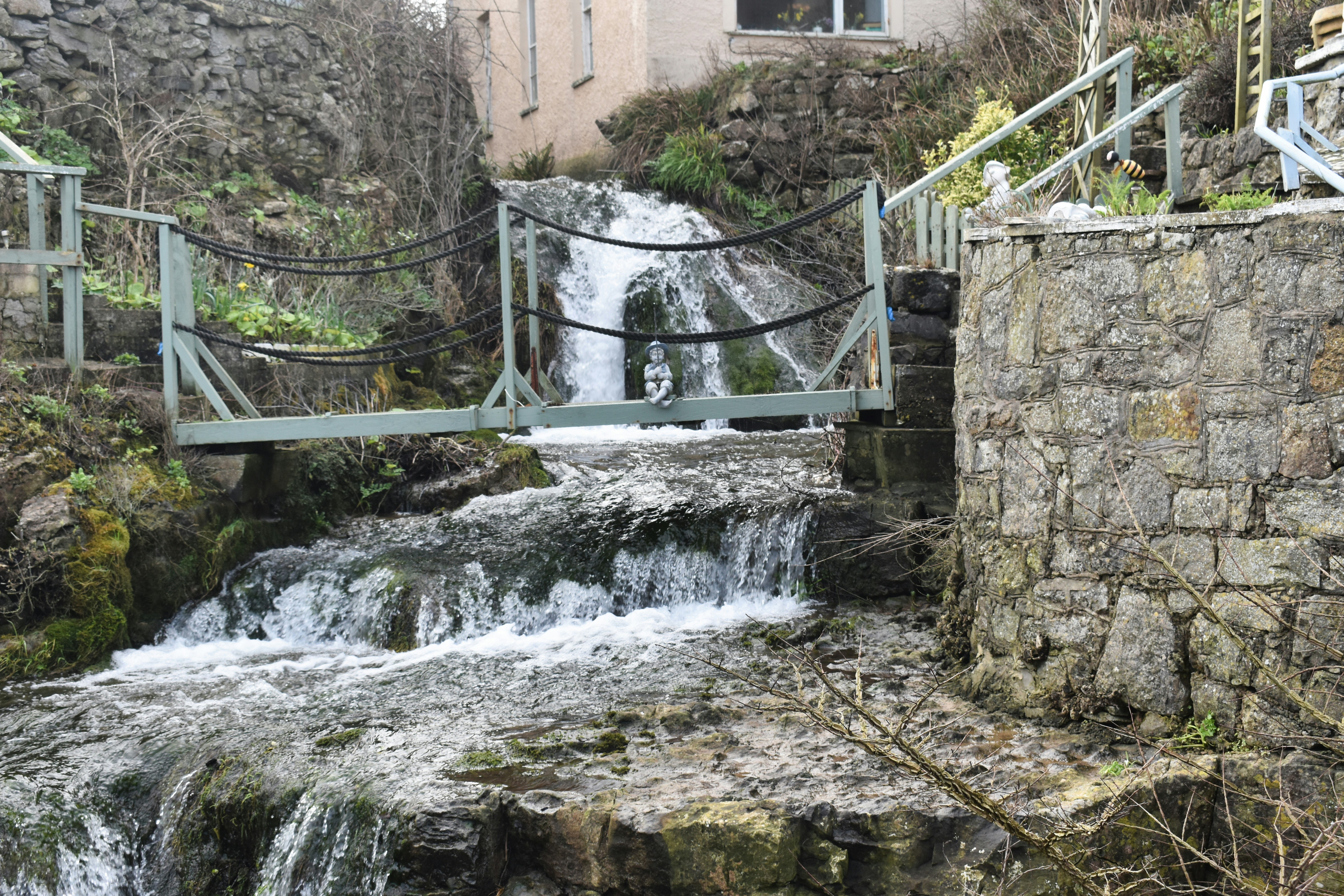 Flowing water cascades over rocks beneath a quaint wooden bridge, framed by lush greenery and a stone wall. A serene rural setting evokes tranquility.
