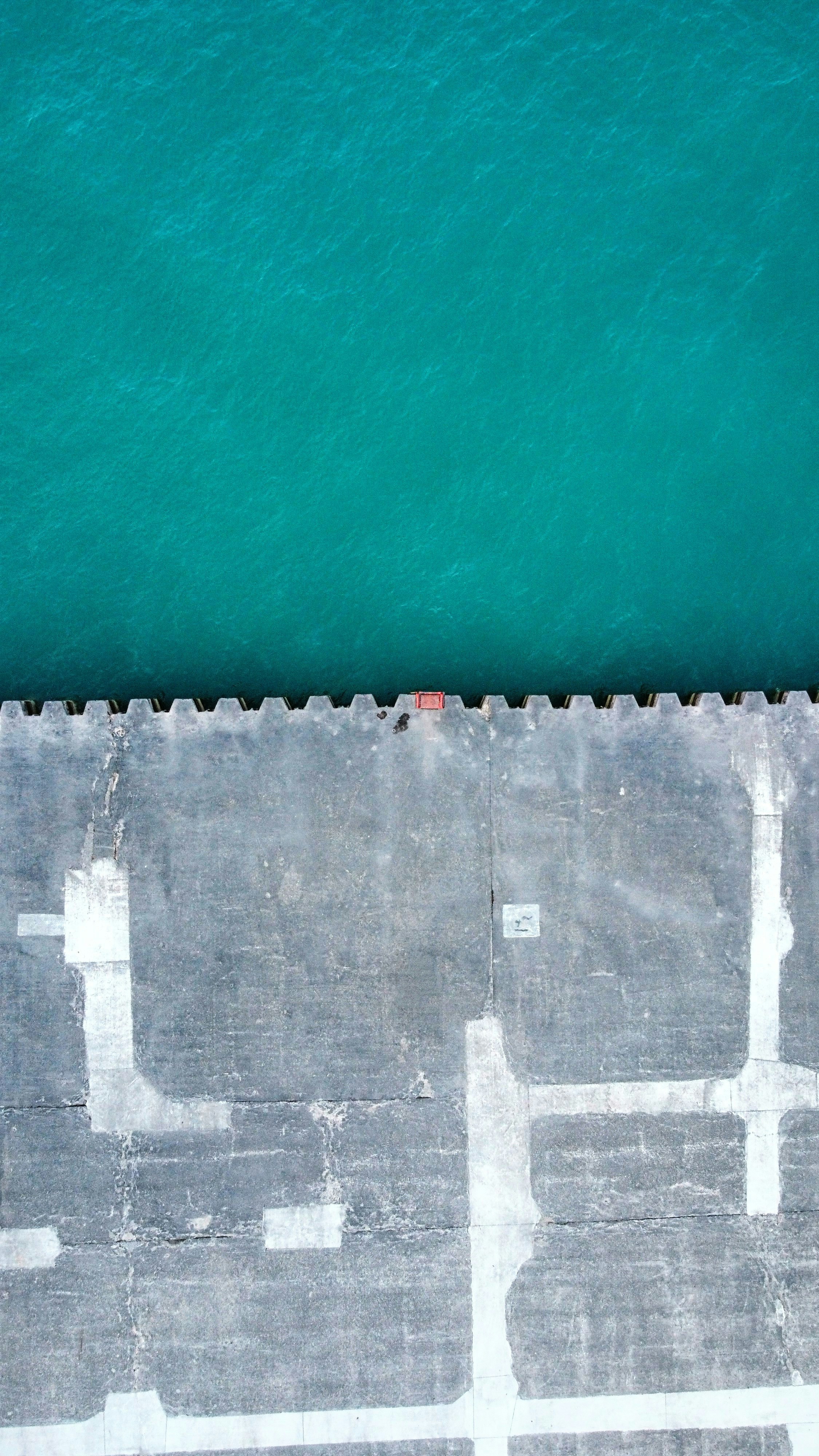 Aerial view of a weathered concrete pier edge meeting turquoise water, with a small red marker at the boundary. The shot emphasizes bold color contrast and geometric lines from a drone perspective.